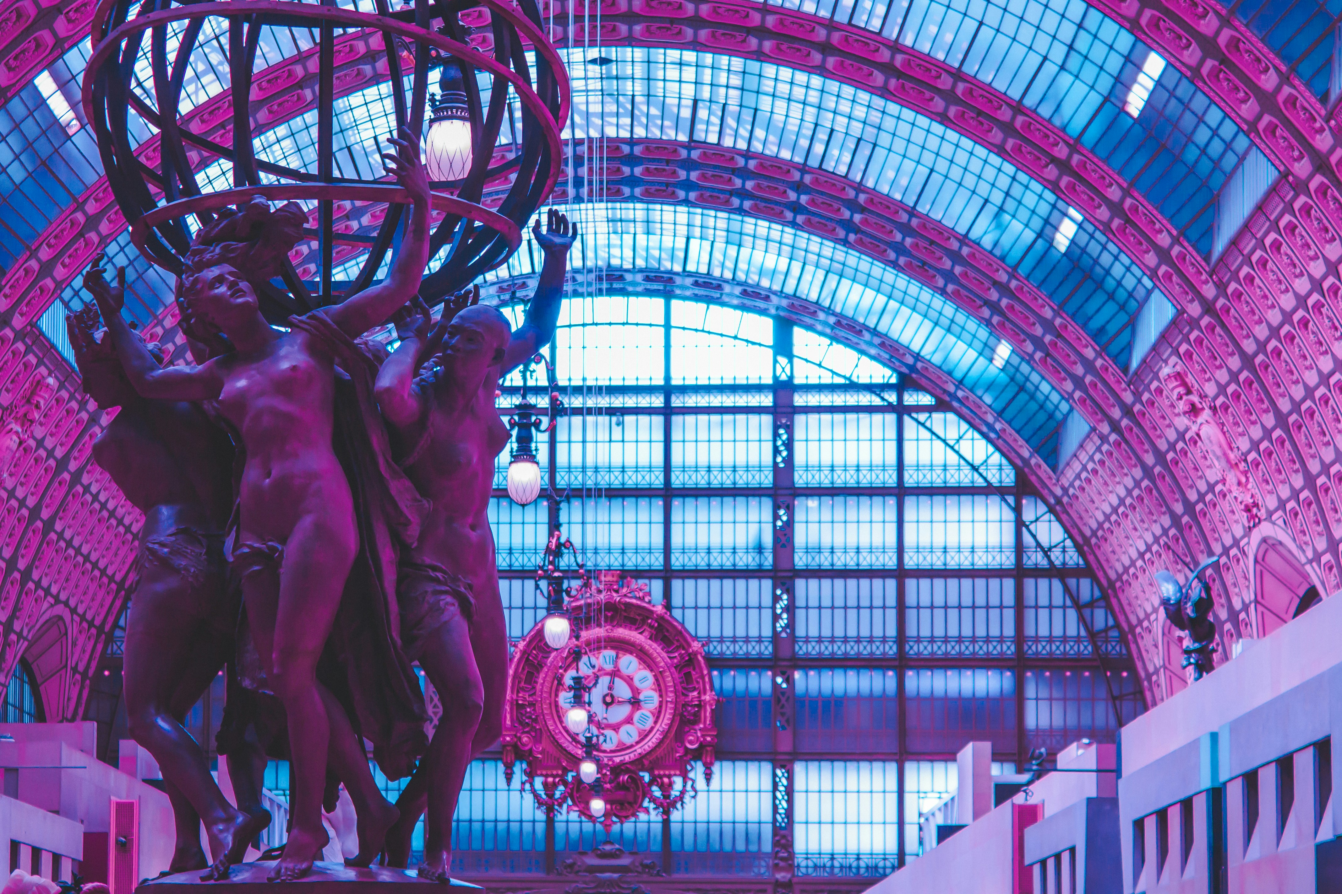 Sculpture under a vibrant pink-lit glass roof with an ornate clock in the background.