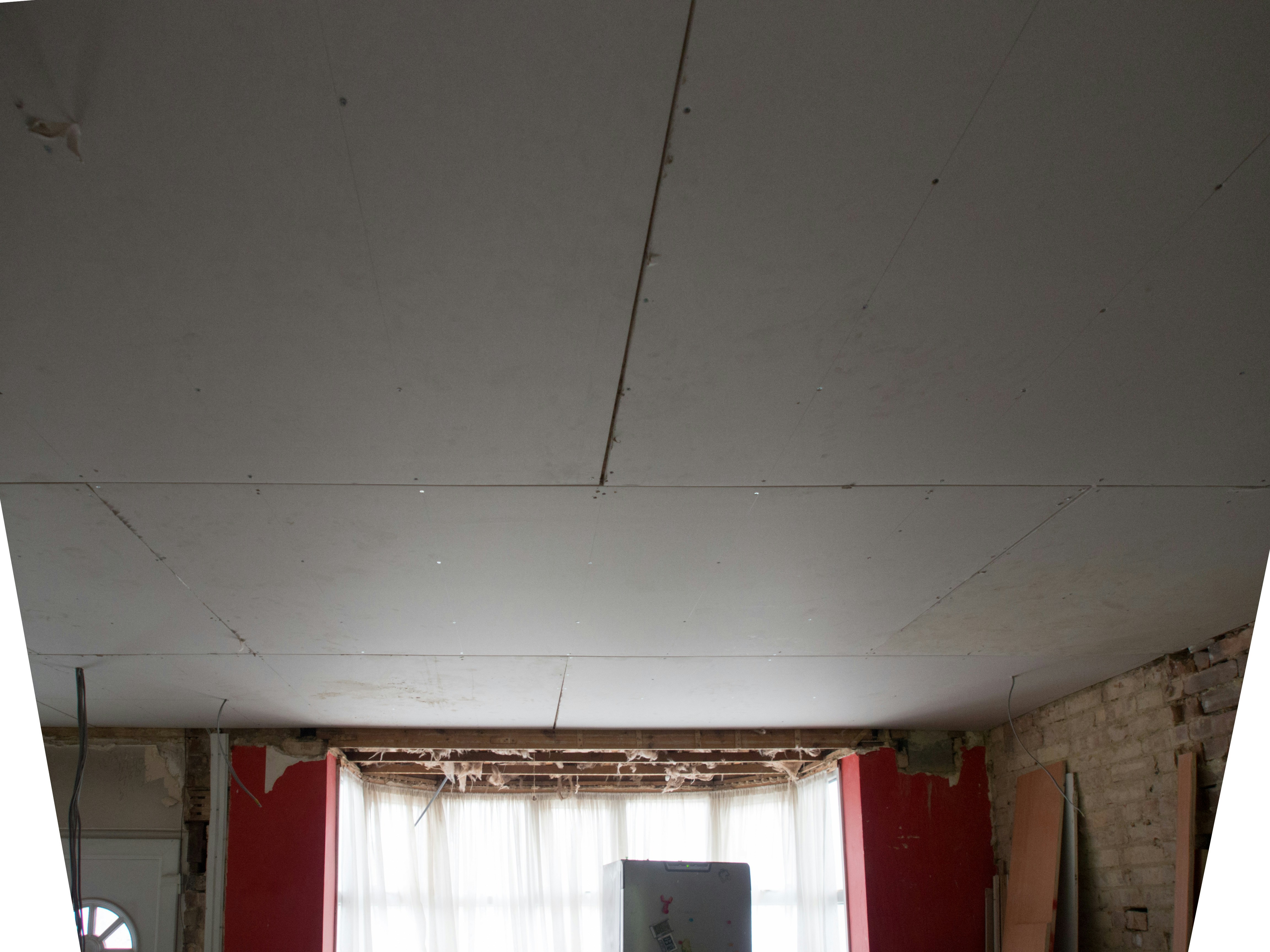 Interior view of a dilapidated room with a cracked ceiling and exposed walls, featuring a window with sheer curtains. The scene conveys a sense of neglect and decay.