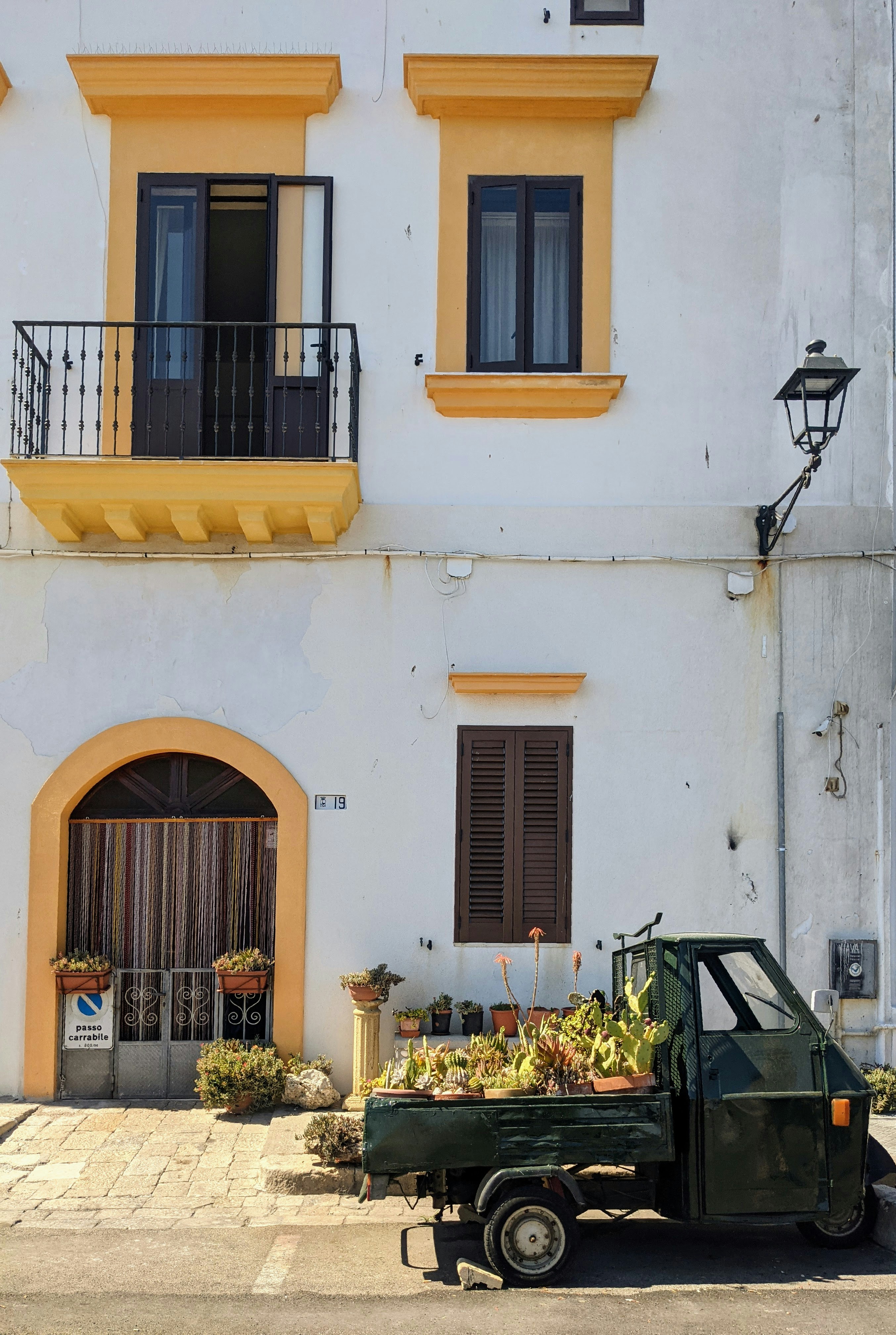 Green truck filled with plants parked in front of a white building with yellow-trimmed windows and balcony.
