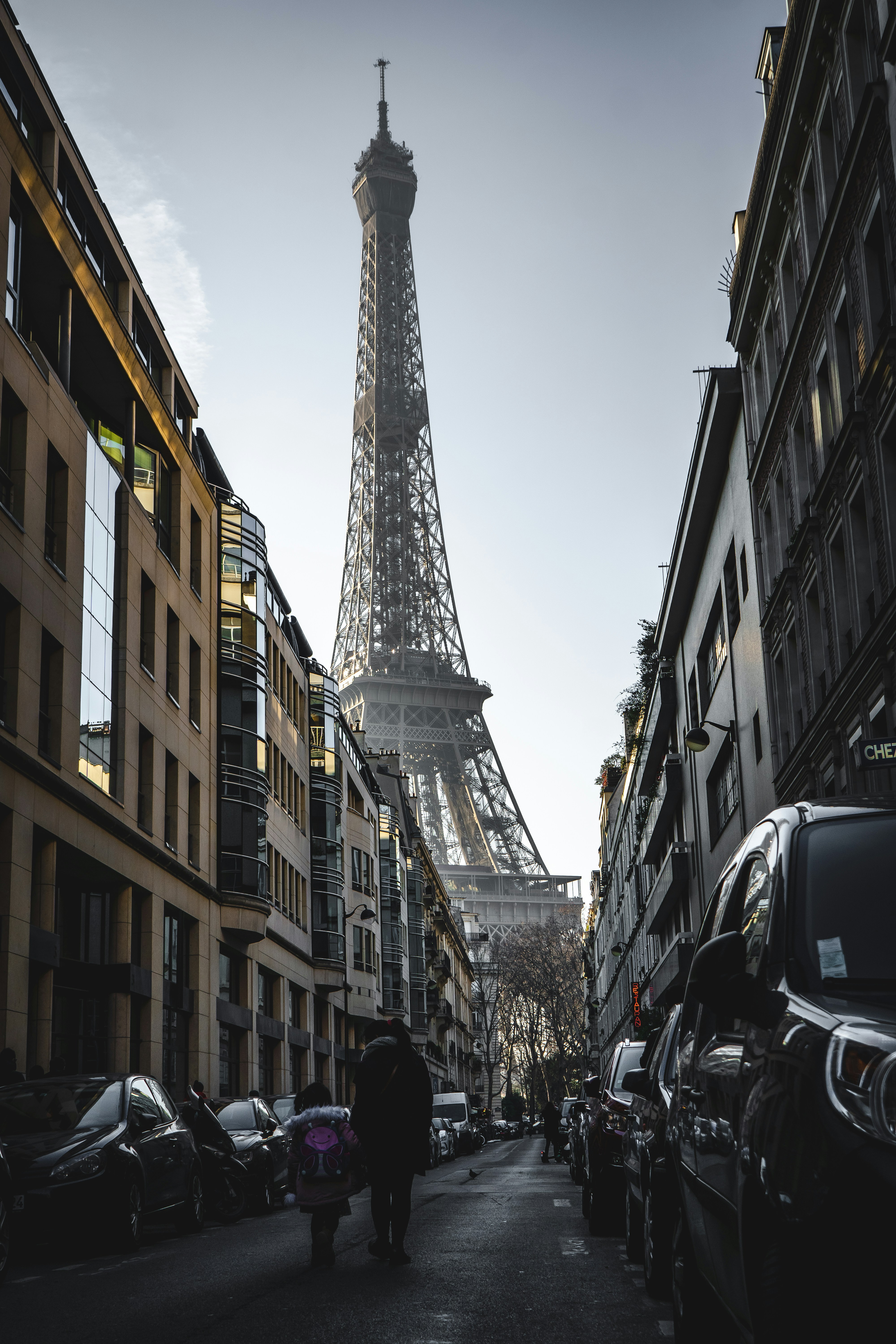 The Eiffel Tower rises majestically above a bustling Parisian street, framed by modern buildings and parked cars. A couple strolls hand-in-hand, adding a touch of life to the scene.