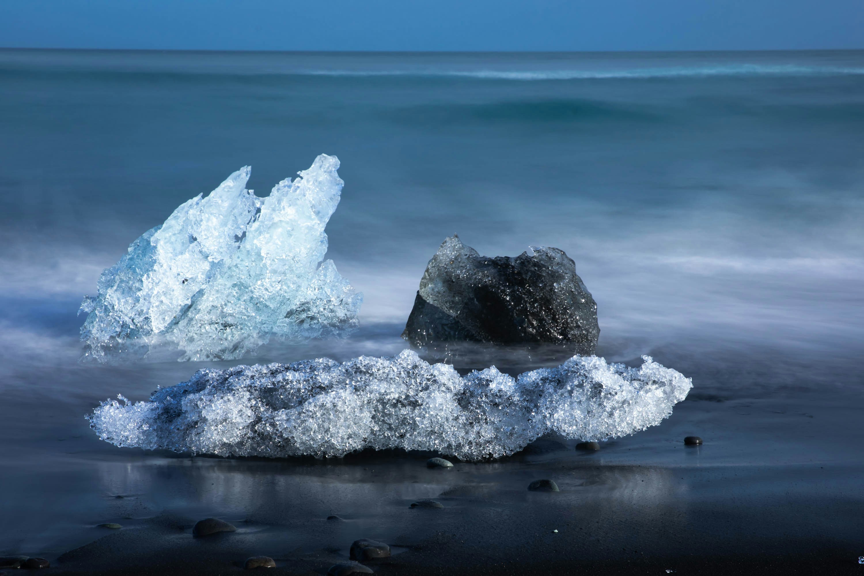 Three ice formations resting on a dark beach, contrasting with the smooth, rolling waves of the ocean. The scene captures the interplay of light and texture in a serene coastal setting.