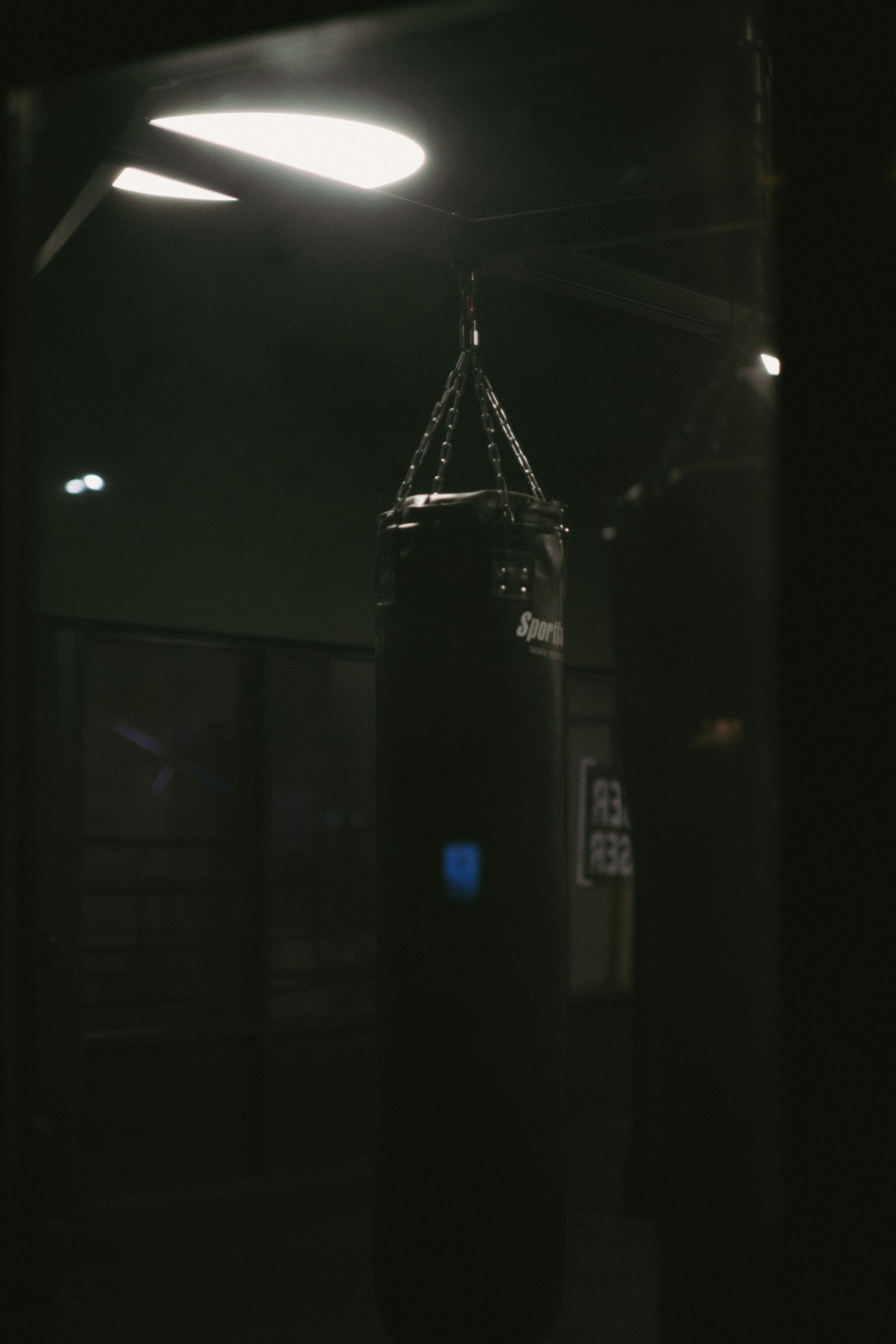 A solitary black punching bag hangs in a dimly lit boxing gym, surrounded by shadows and hints of light. The atmosphere conveys a sense of determination and focus.