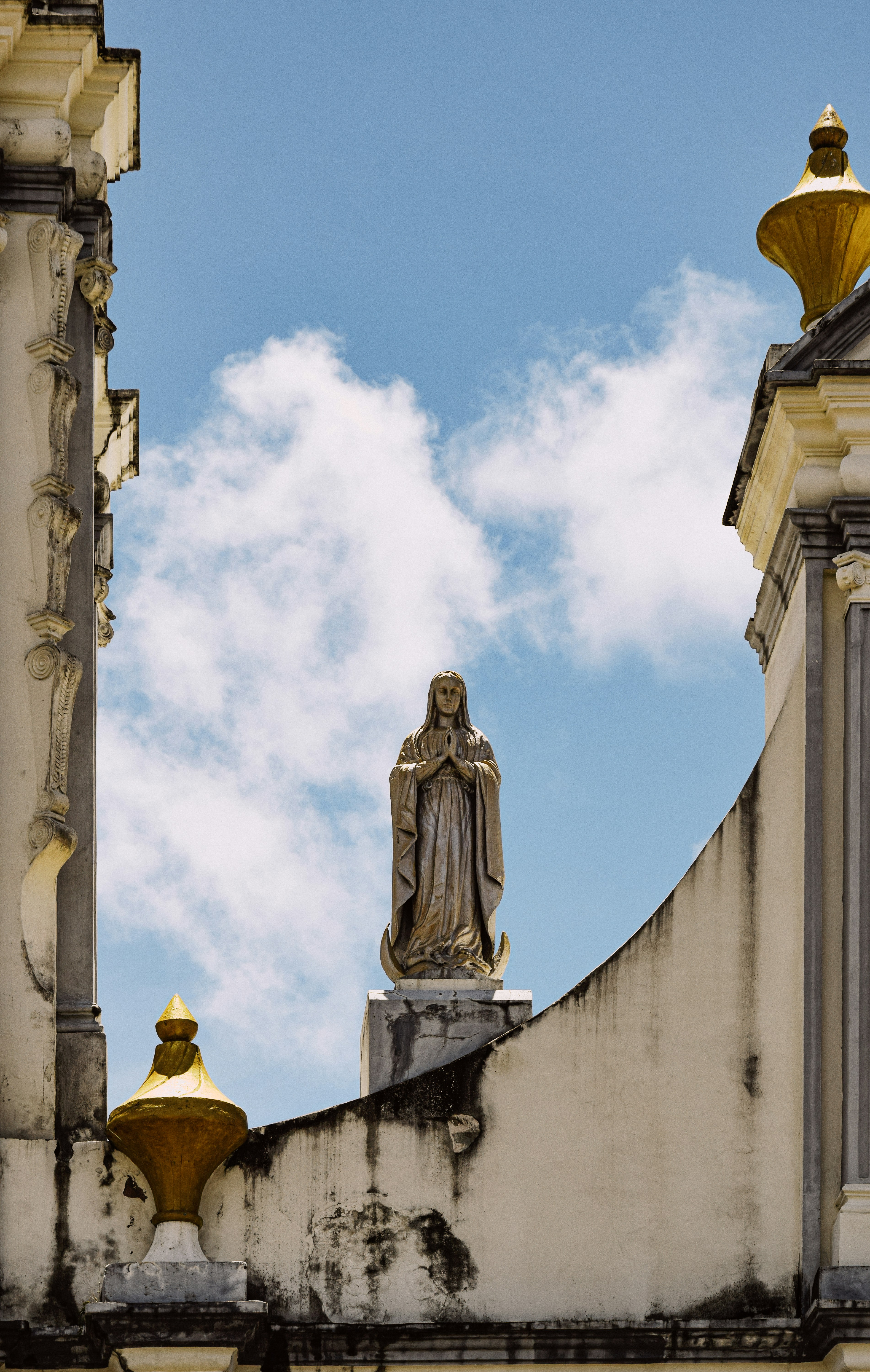 A statue of a figure stands atop a weathered wall, framed by a bright blue sky and wispy clouds, evoking a sense of serenity and history.