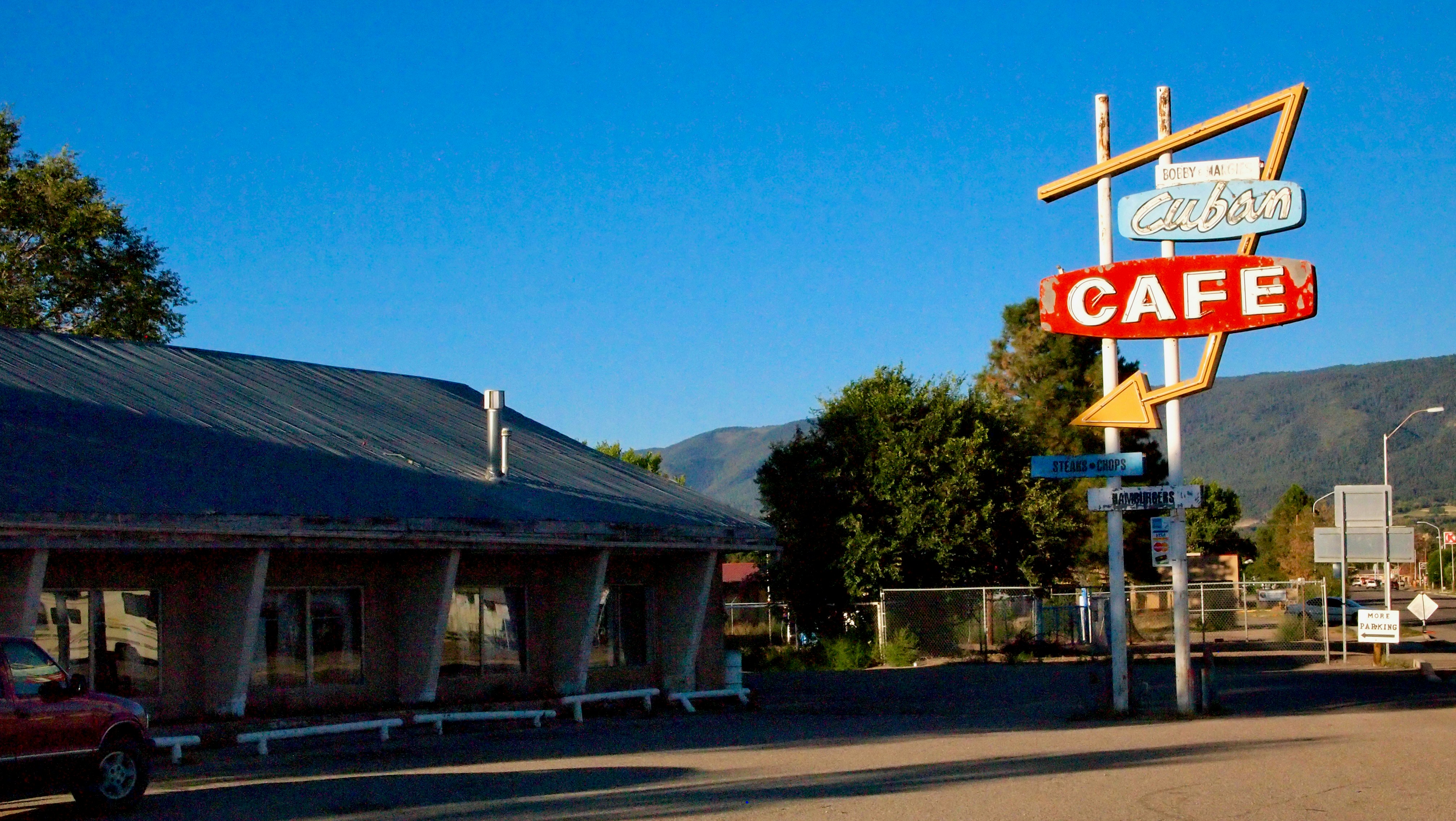 Vintage café sign with a bold red 'CAFÉ' against a clear blue sky, surrounded by trees and mountains.
