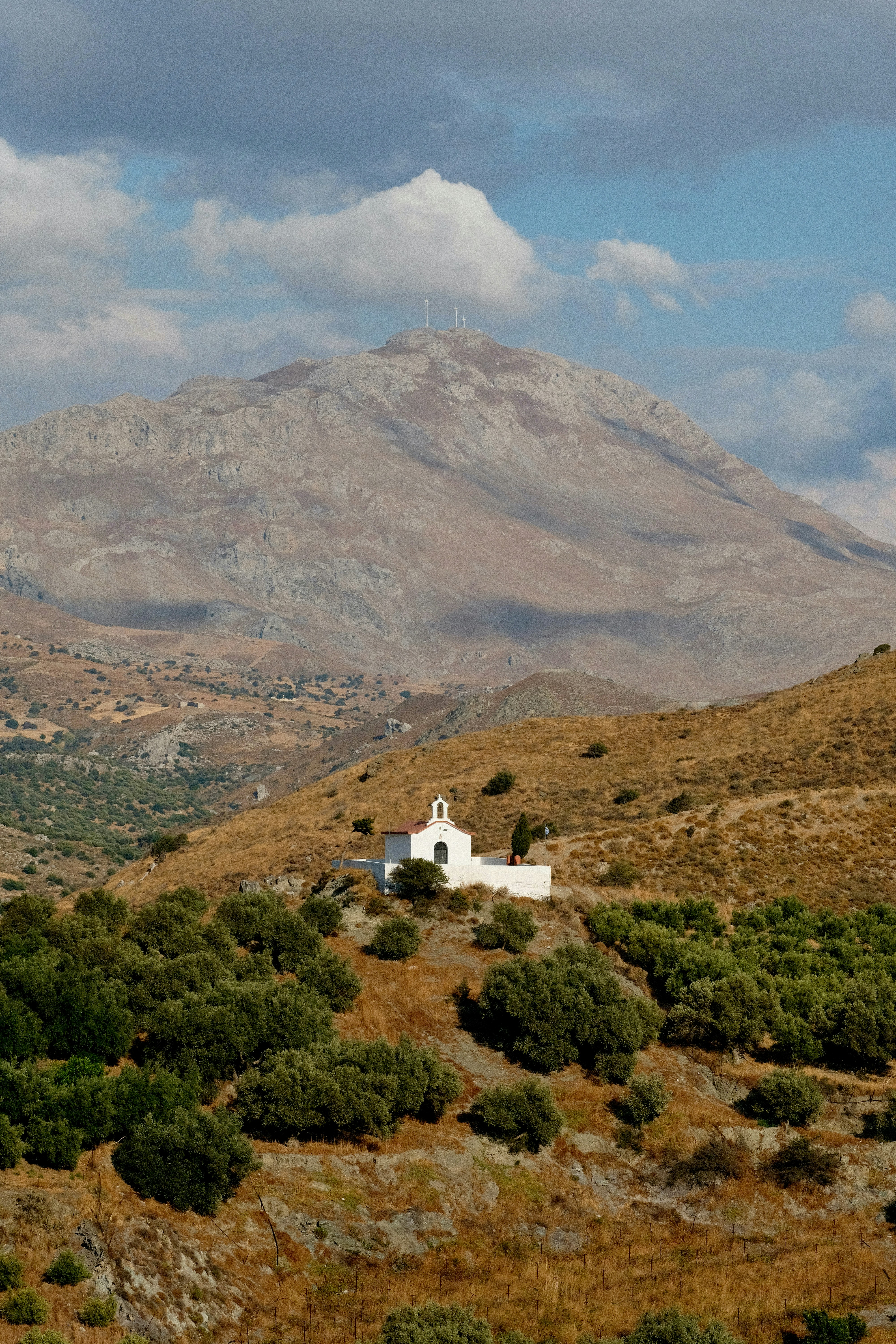 A quaint white chapel nestled among olive trees, with a majestic mountain backdrop under a partly cloudy sky.