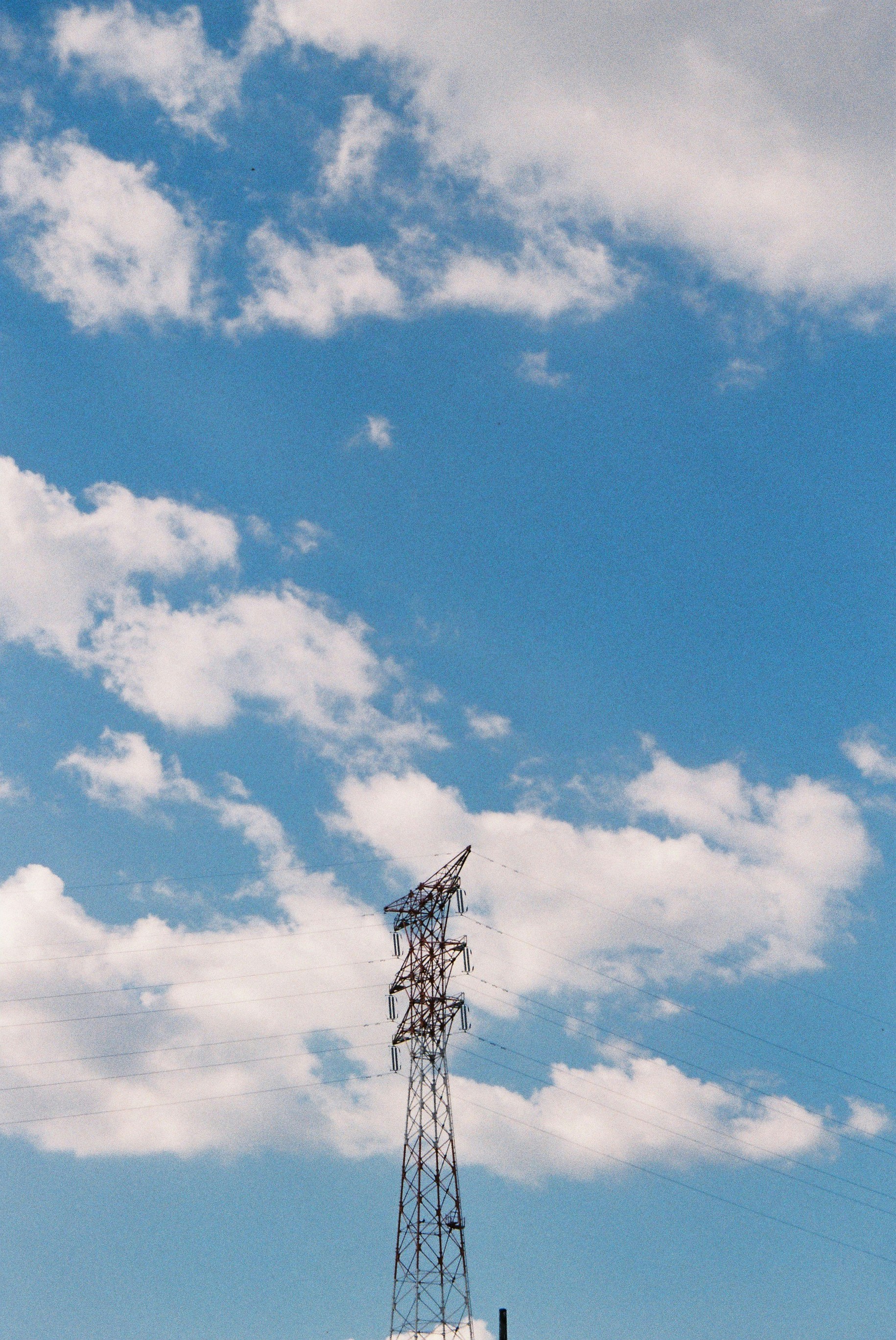 A tall power line tower stands against a backdrop of scattered clouds in a bright blue sky.