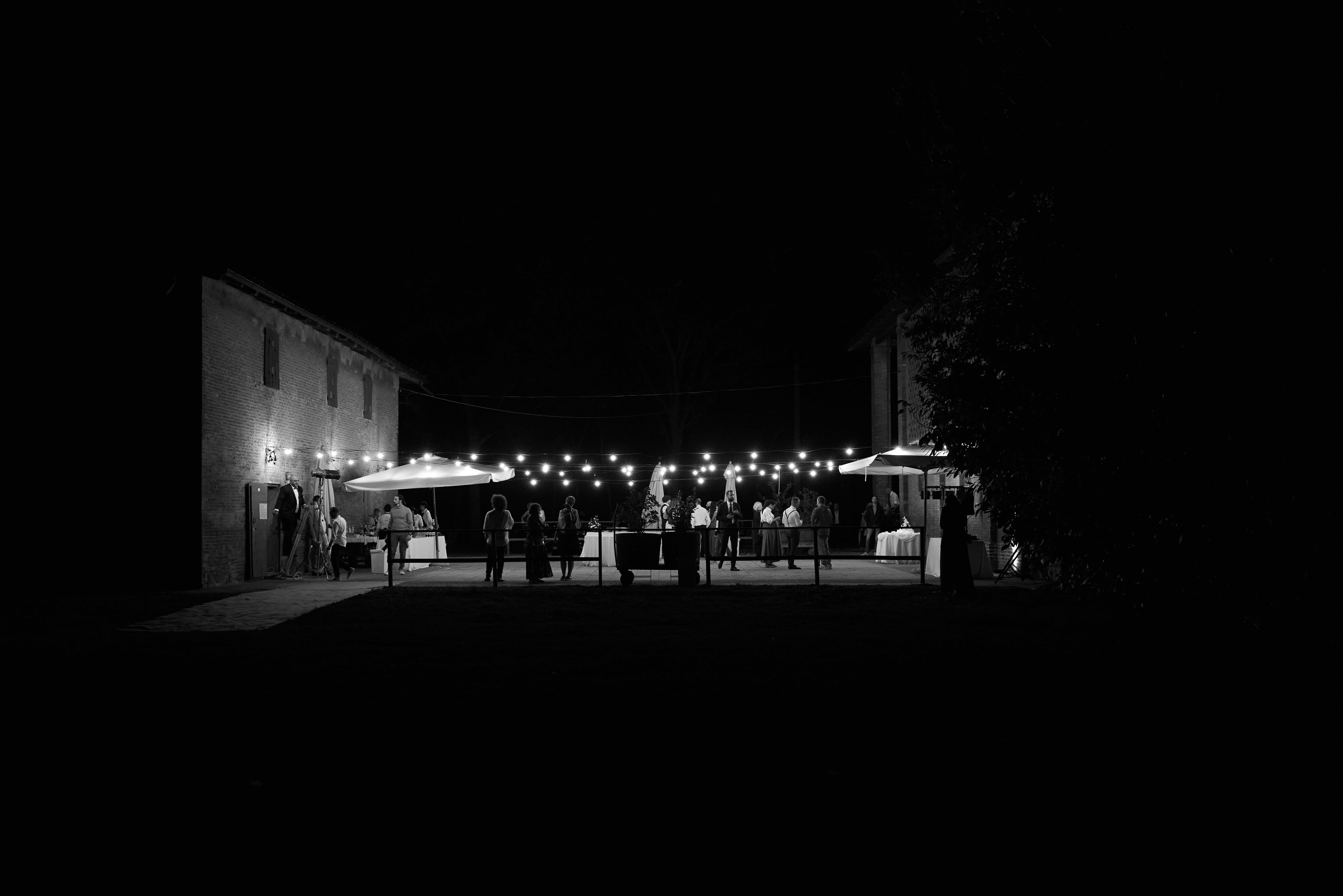 Gathering of people enjoying an outdoor event illuminated by string lights, set against a rustic backdrop at night.