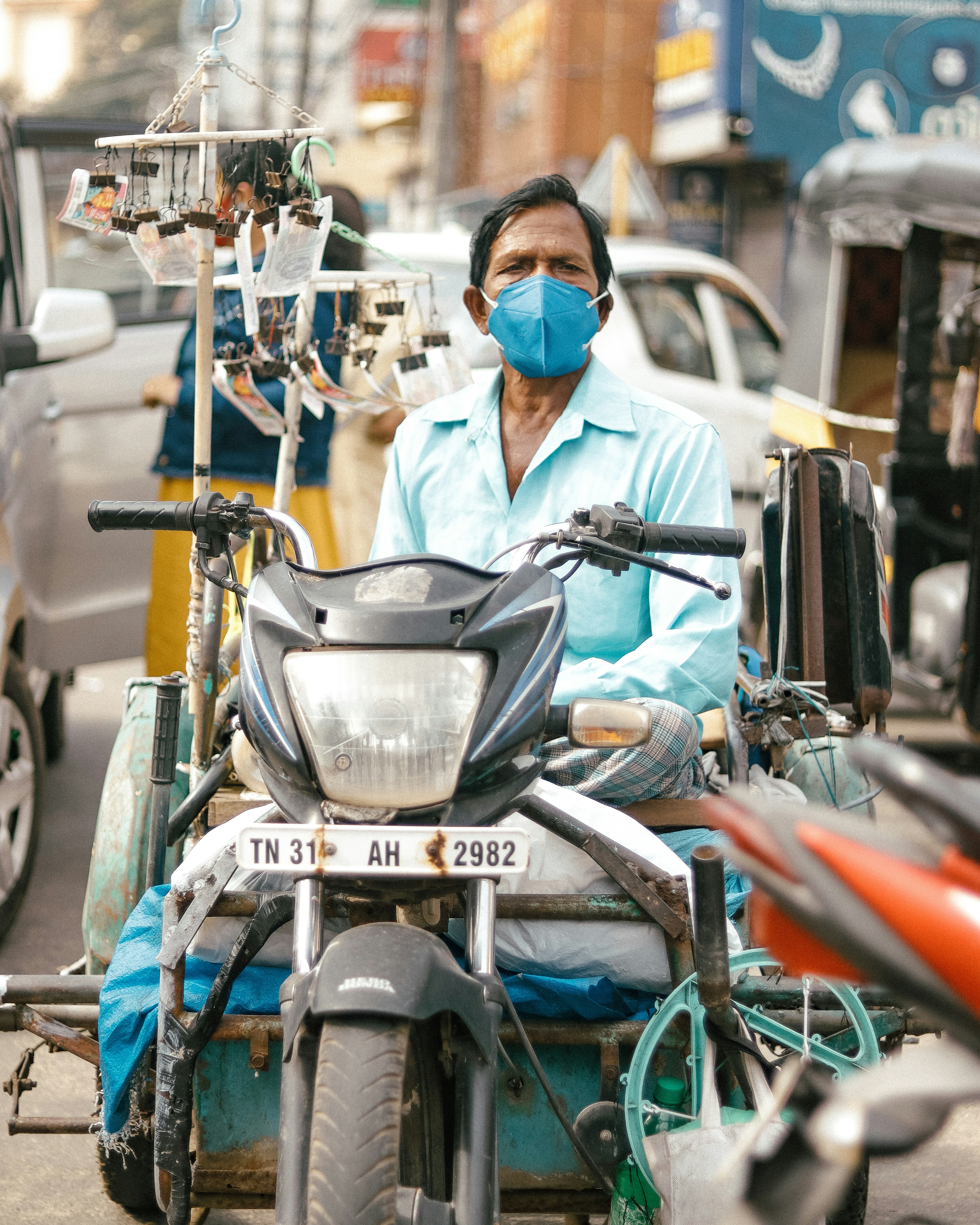 A street vendor wearing a blue mask sits on a motorcycle, surrounded by various goods for sale in a bustling urban setting.