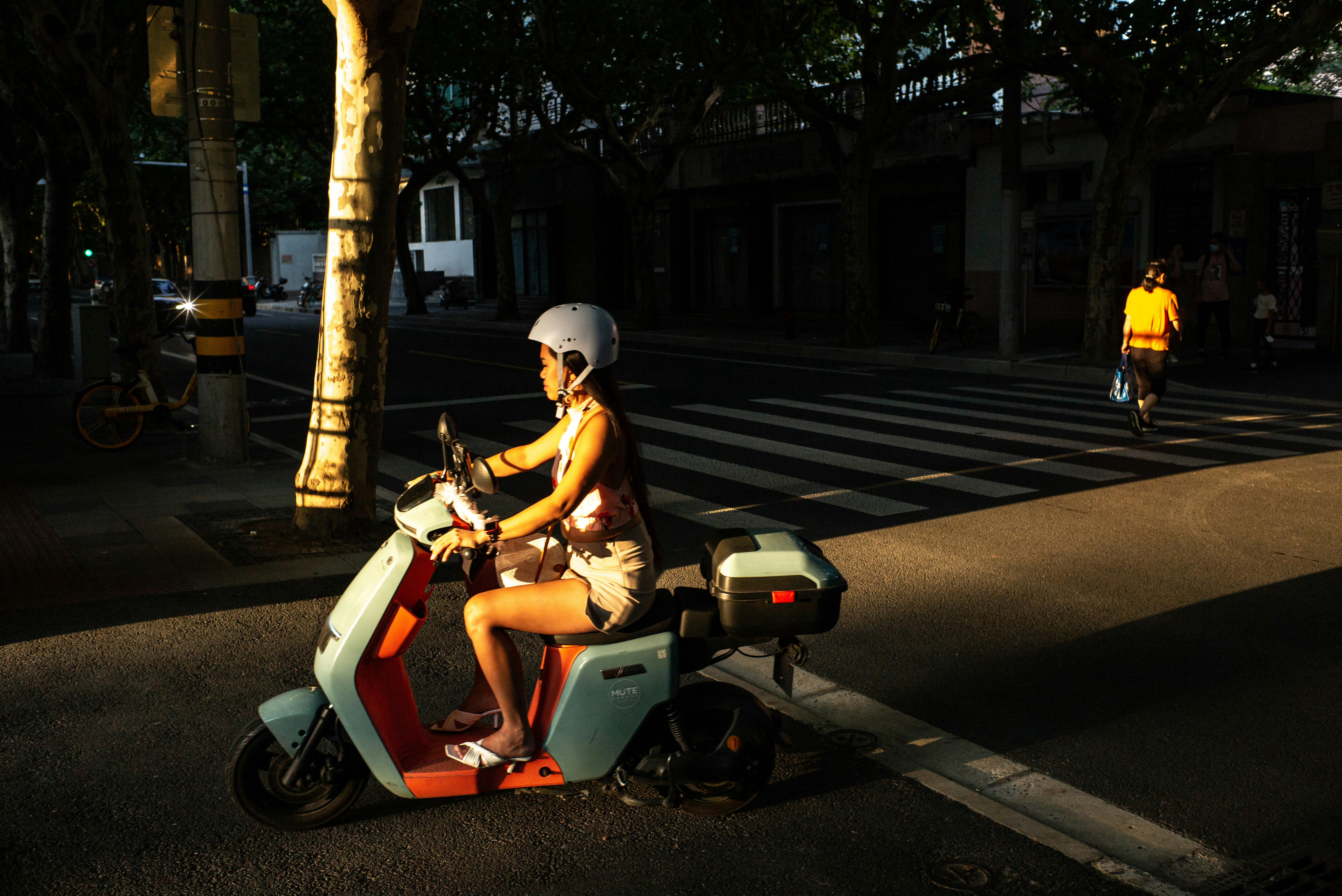 A woman riding a scooter bathed in warm sunlight, navigating a quiet city street with shadows and vibrant colors. The scene captures the essence of urban life at dusk.