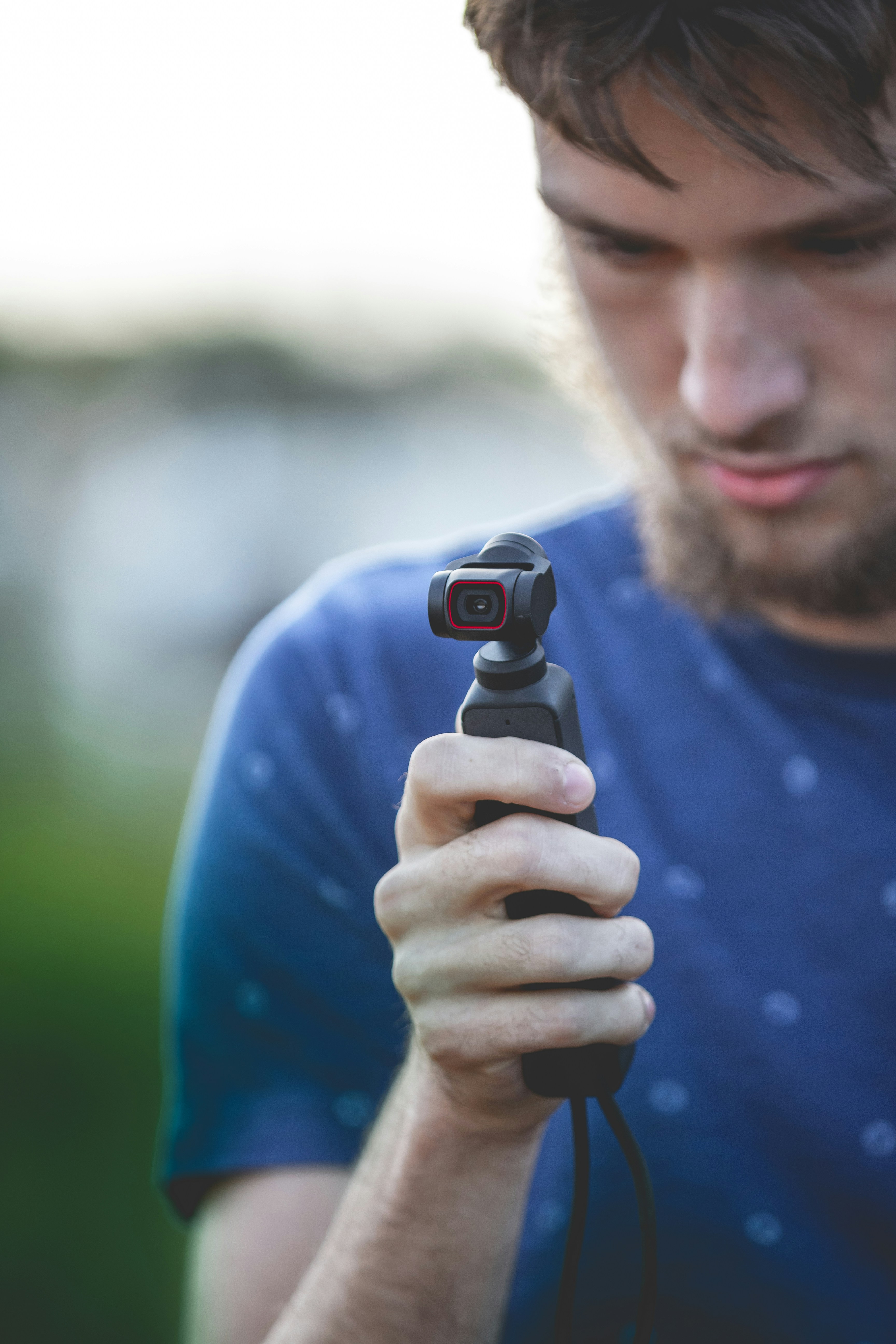 Young man focused on a handheld camera stabilizer, preparing for a shoot in a blurred outdoor setting.