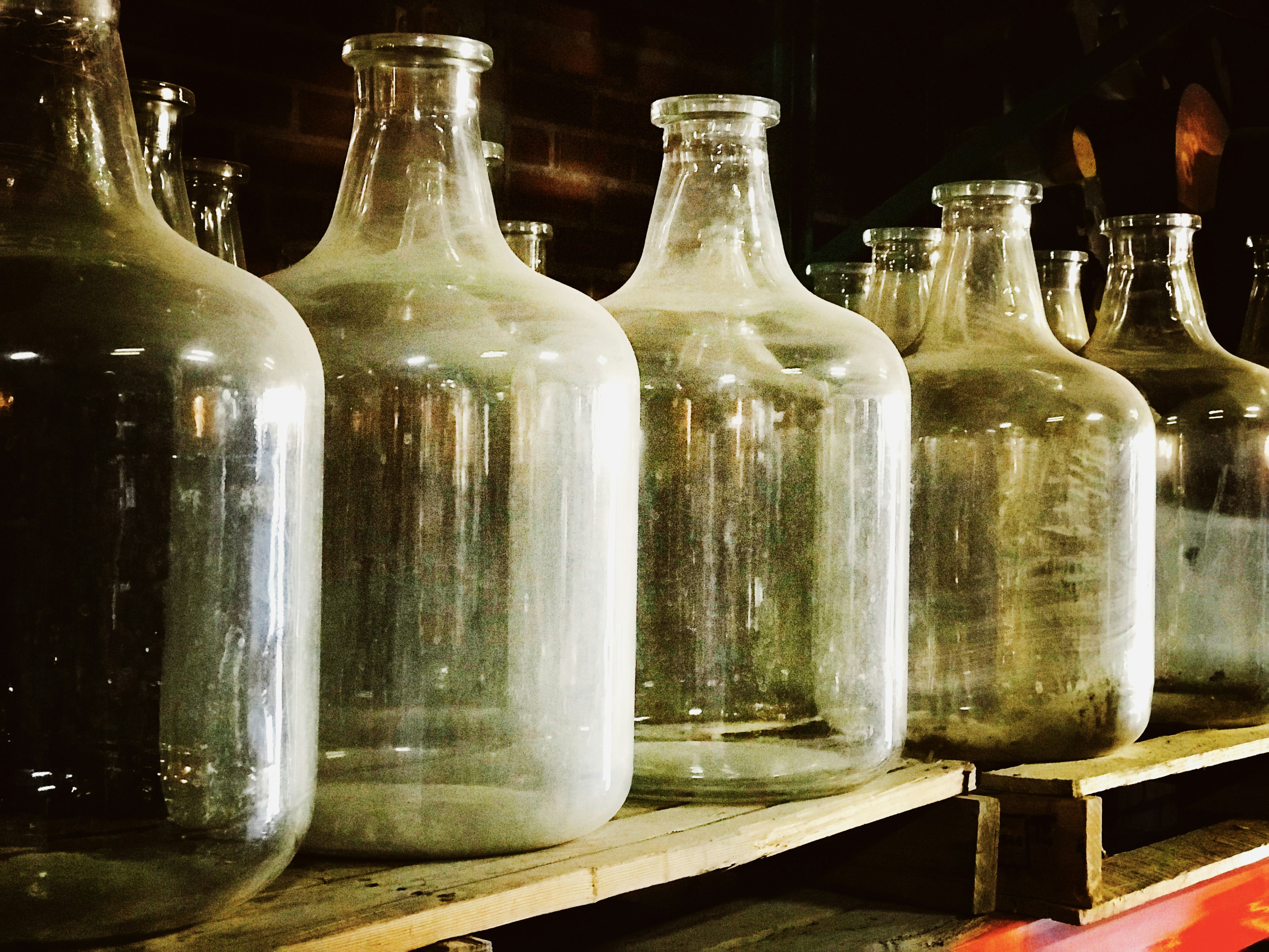 Collection of clear glass bottles arranged on wooden shelves, showcasing their unique shapes and reflections in a dimly lit environment.