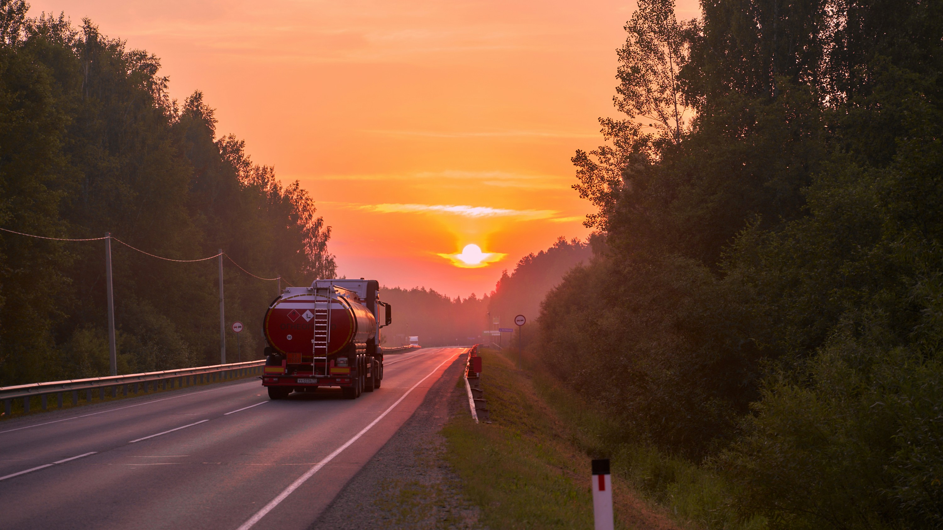 Truck traveling down a forest-lined highway at sunset with vibrant orange sky.