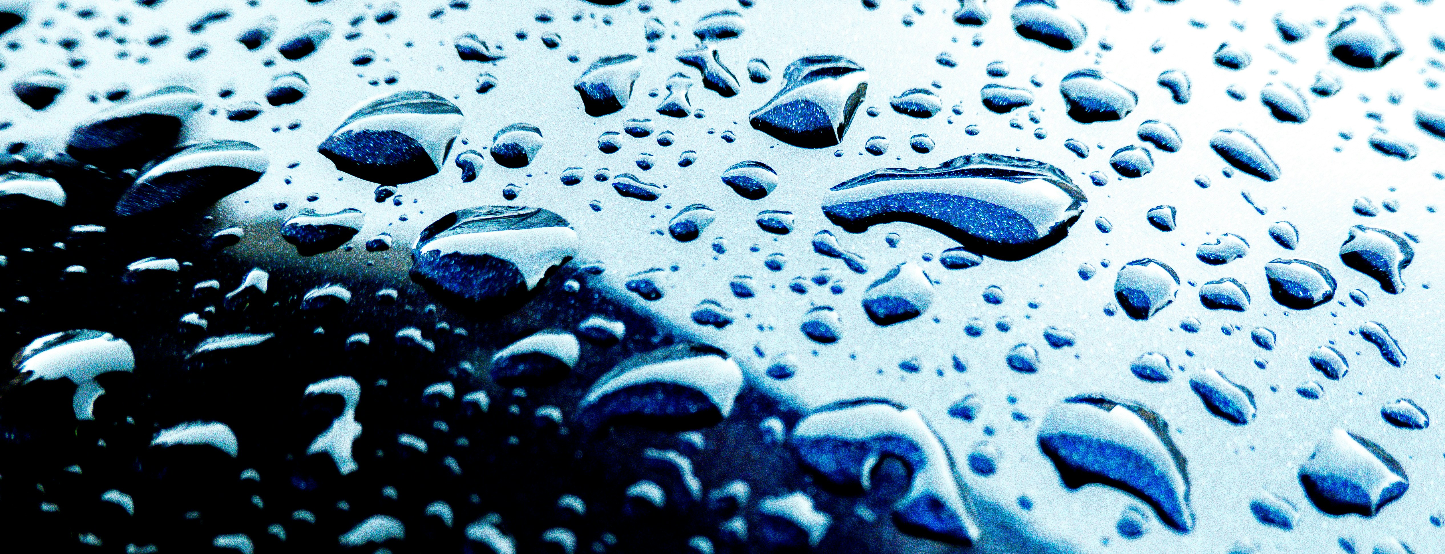 Close-up of water droplets resting on a smooth surface, showcasing intricate reflections and textures. The image highlights the interplay of light and water.