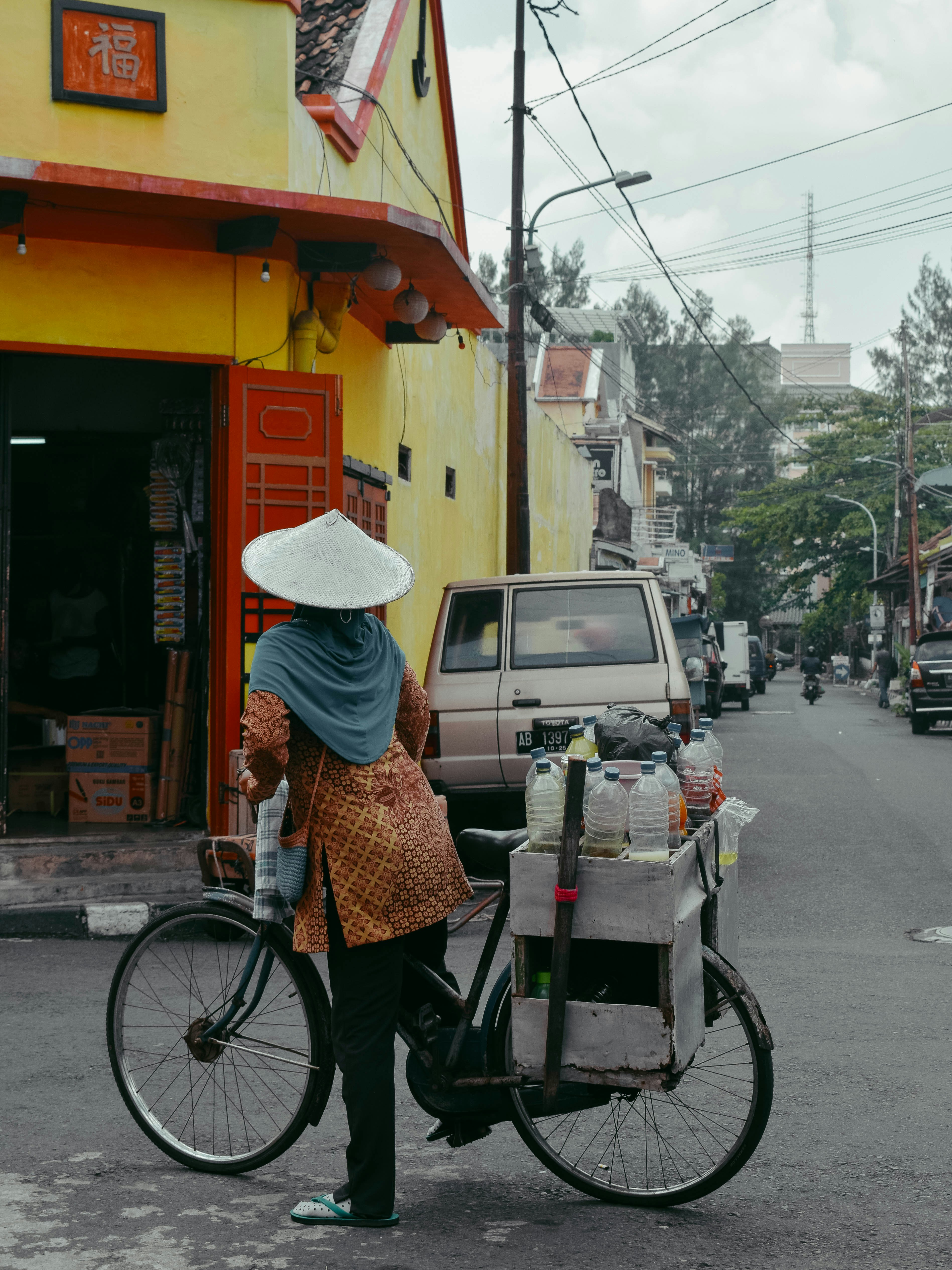 A vendor in traditional attire rides a bicycle loaded with bottled goods, set against a vibrant yellow building in an urban street scene.