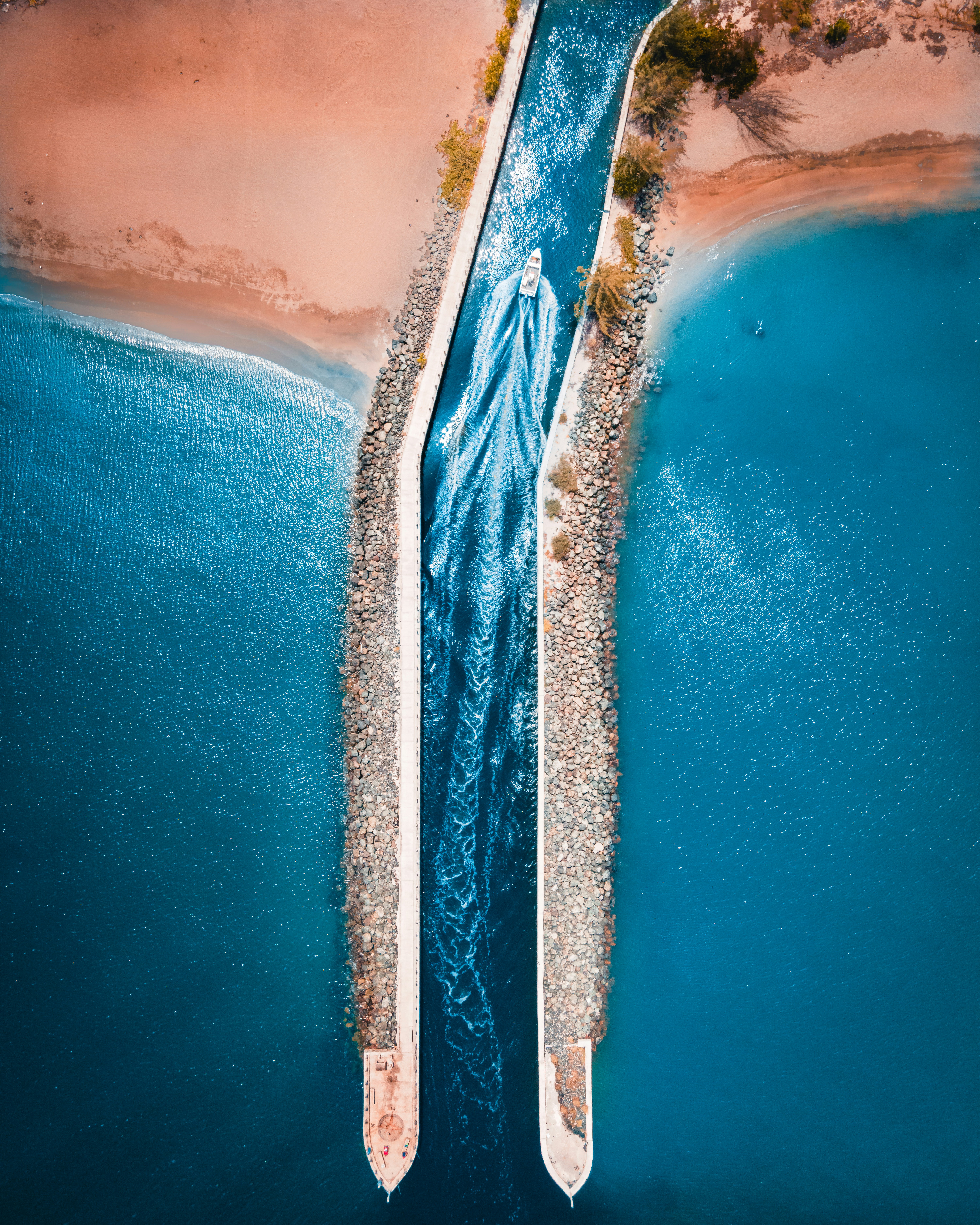 Aerial view of a narrow waterway flanked by sandy shores and rocky barriers, with a boat creating ripples as it moves through the channel.