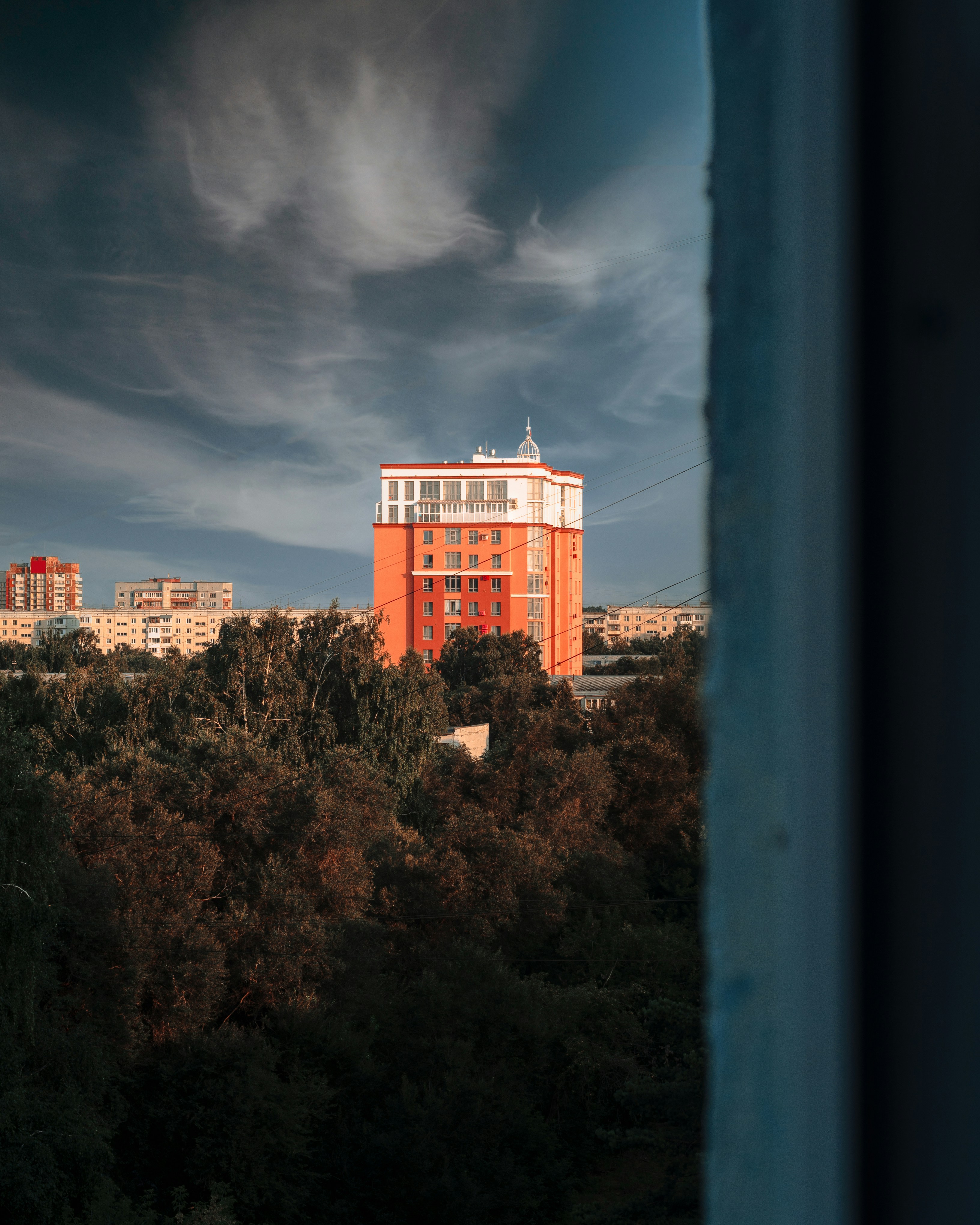 A striking orange building stands amidst lush greenery and urban architecture, framed by a dramatic sky. The scene captures the blend of nature and city life.
