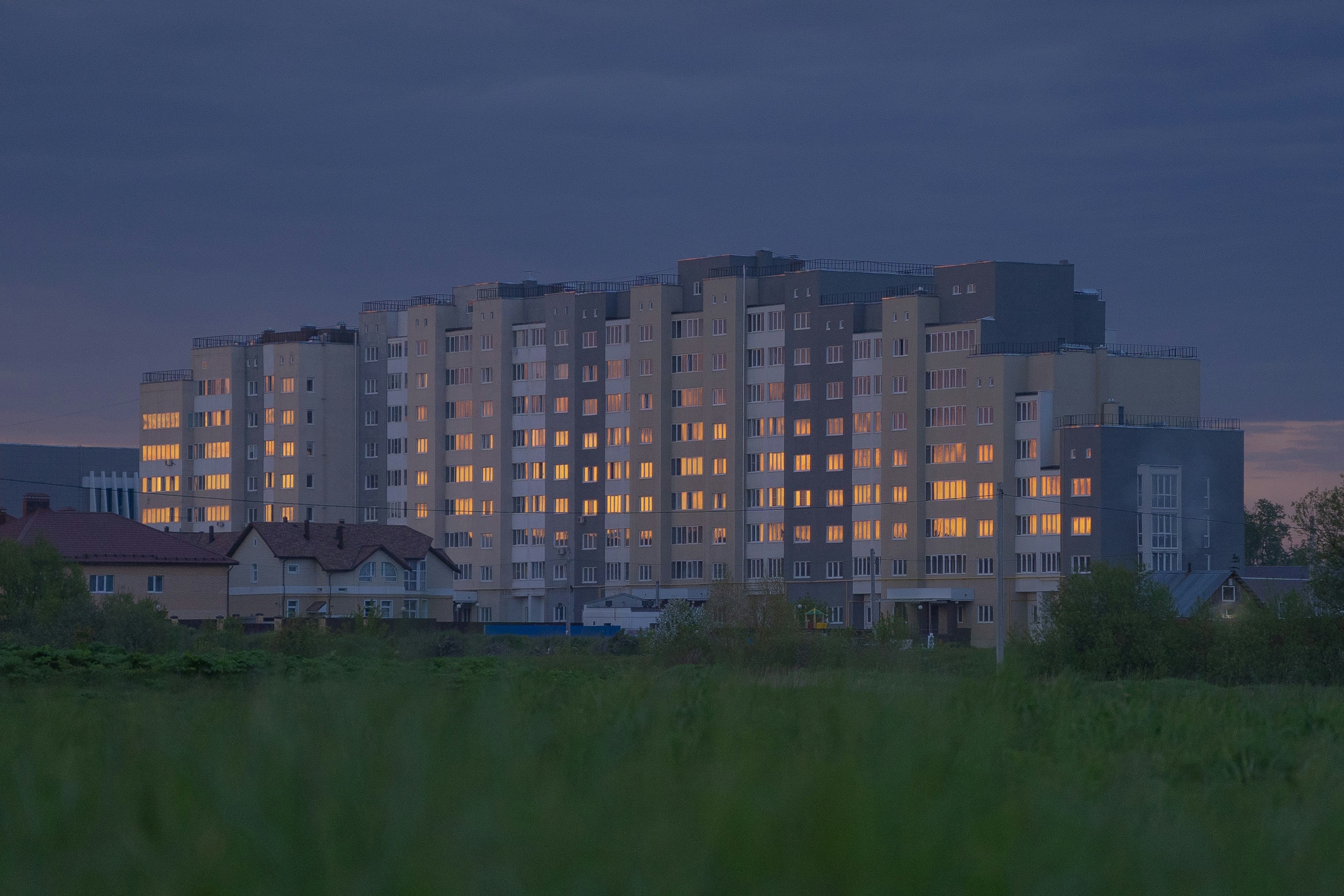 Modern apartment building illuminated by warm light from windows, contrasting against a twilight sky. The surrounding landscape features grassy fields and distant houses.