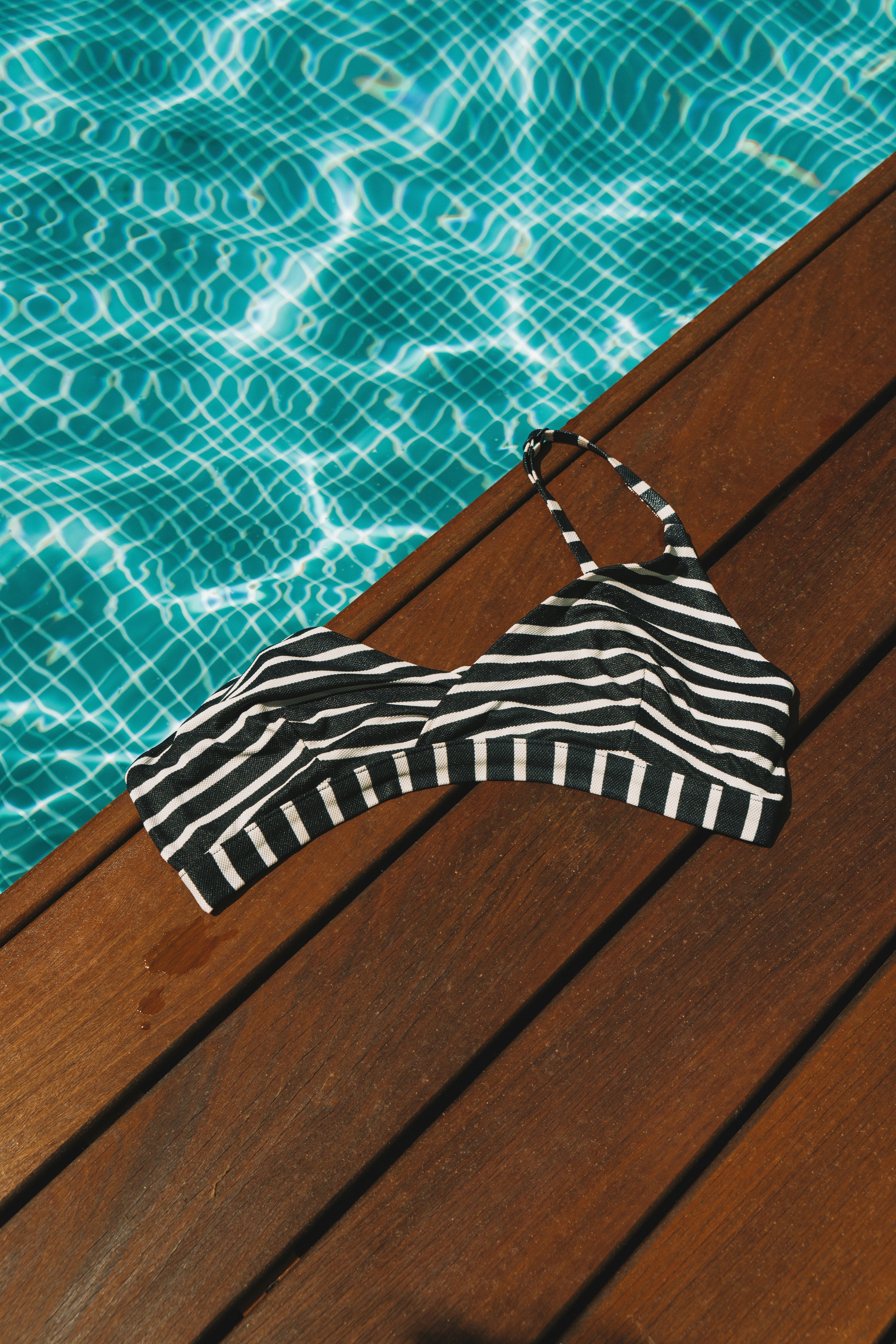 Black and white striped bikini top resting on wooden deck beside a shimmering pool. The sunlight creates playful reflections in the water.