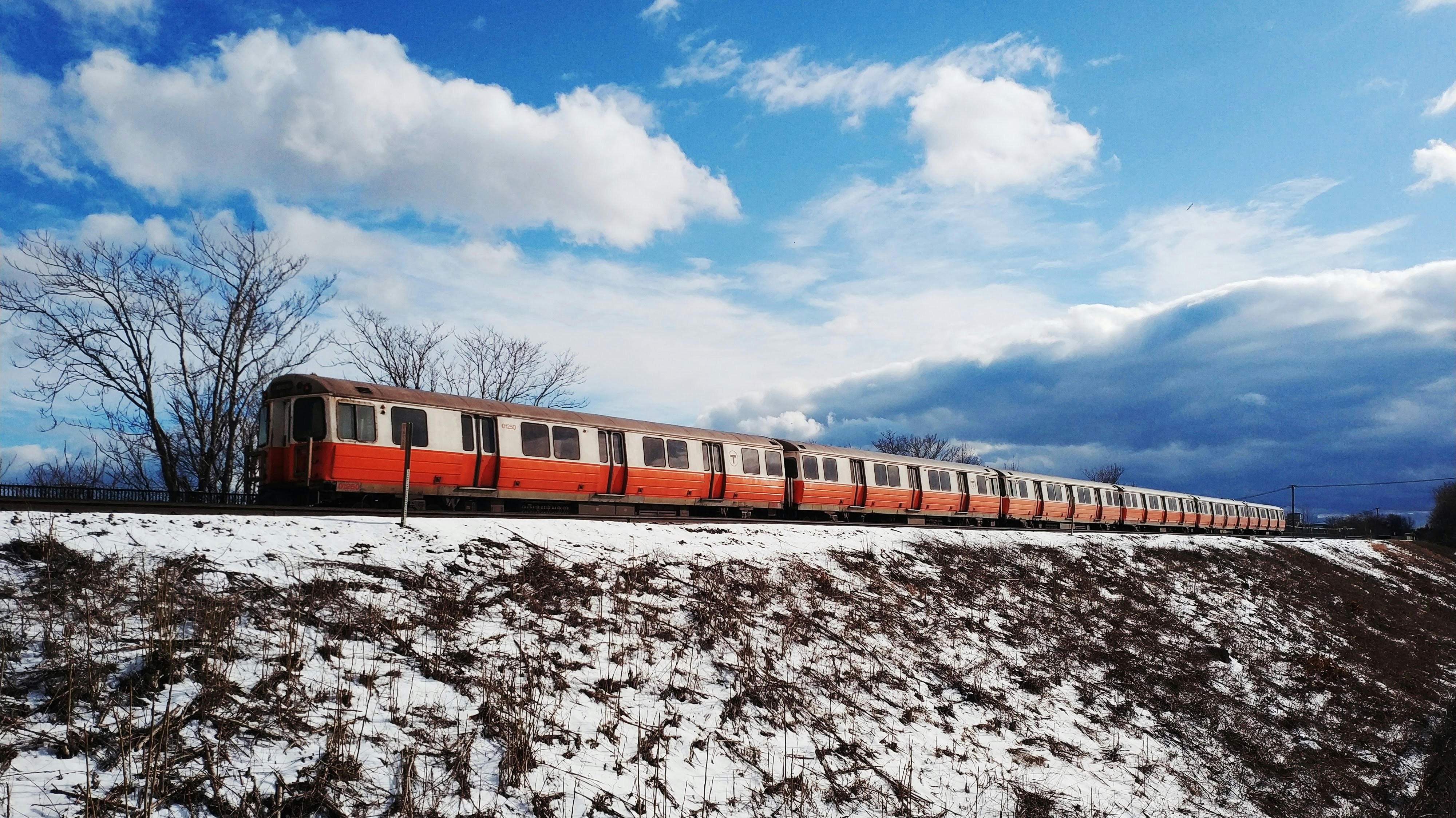 A vibrant orange train glides along a snowy landscape under a dramatic sky, highlighting the contrast between urban transport and nature's tranquility.