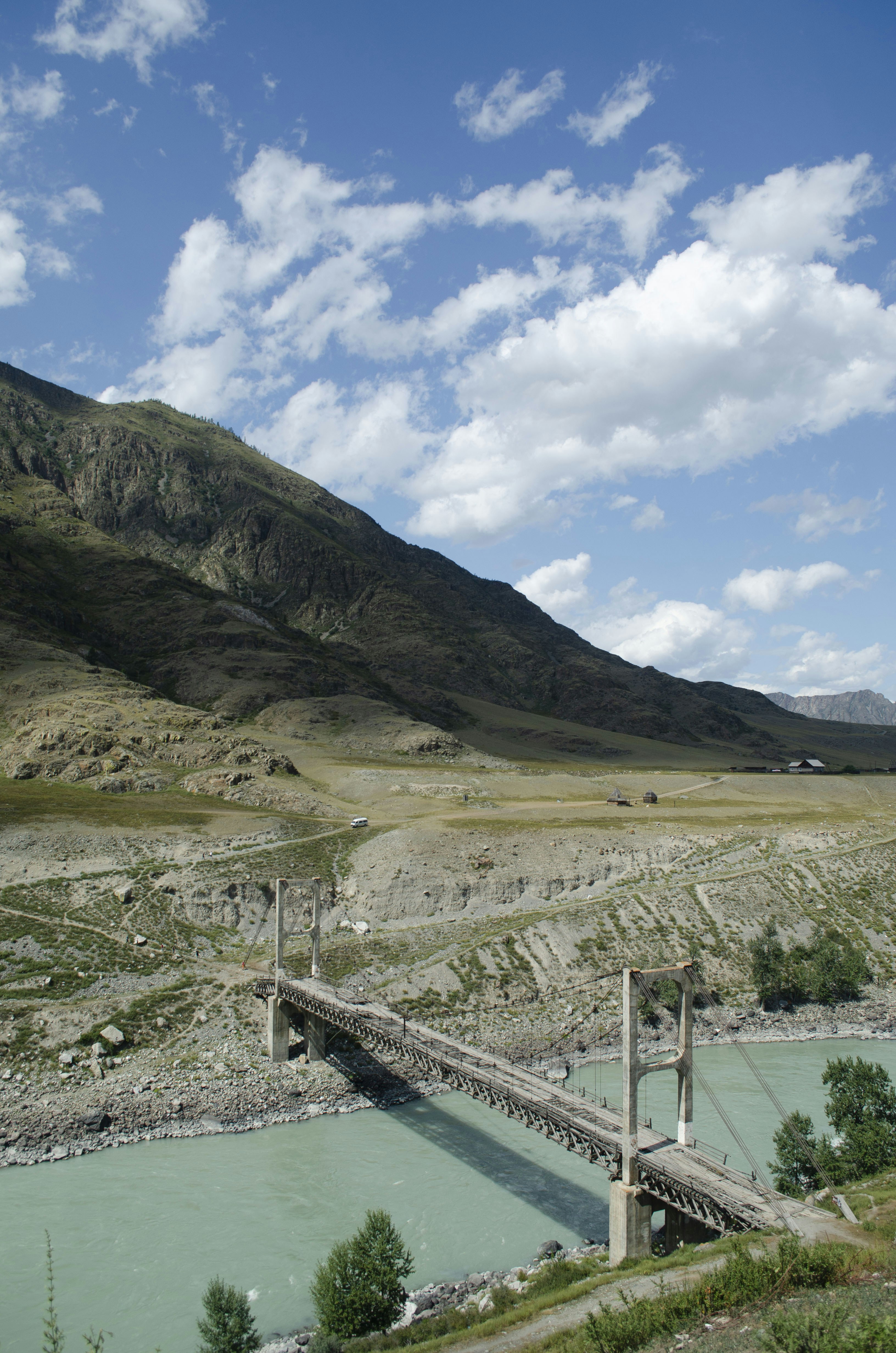 Suspension bridge spanning a turquoise river, framed by rugged mountains and a clear sky. The landscape showcases the harmony between human engineering and natural beauty.