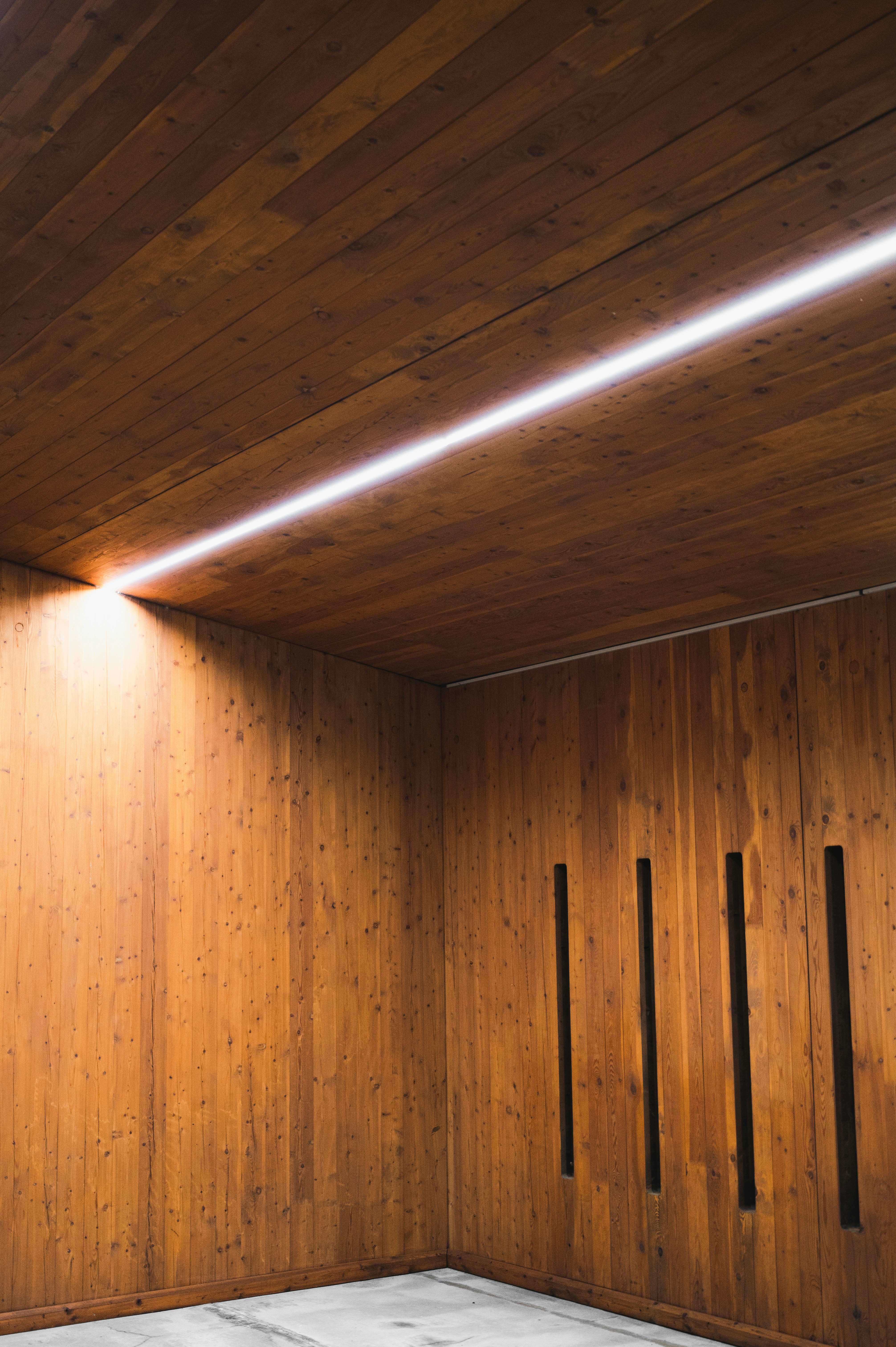 Interior corner showcasing a wooden wall with vertical slits and a linear light fixture above. The warm tones of the wood create a cozy ambiance.