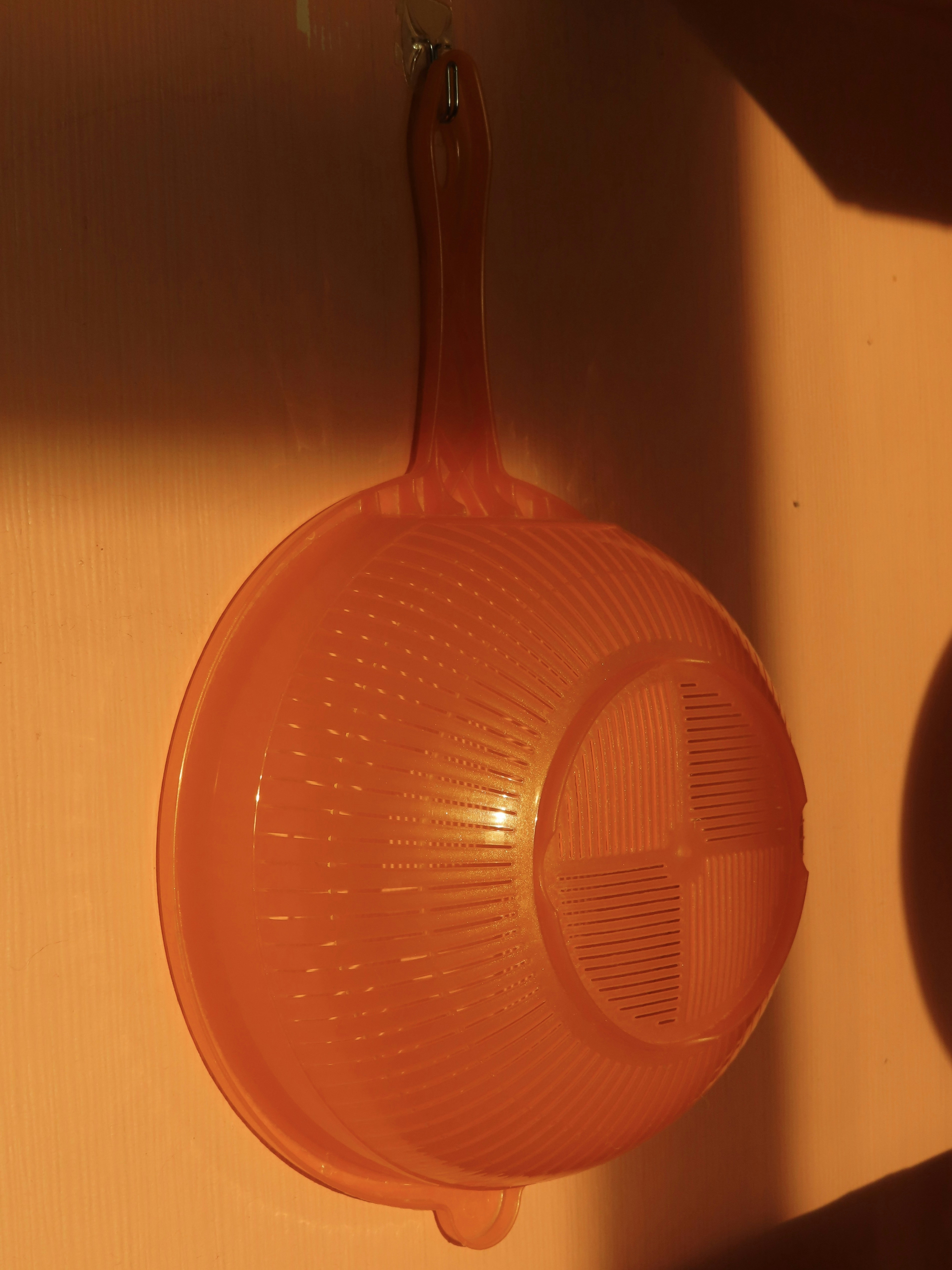 Orange colander hanging against a softly lit wall, showcasing intricate patterns through shadows.