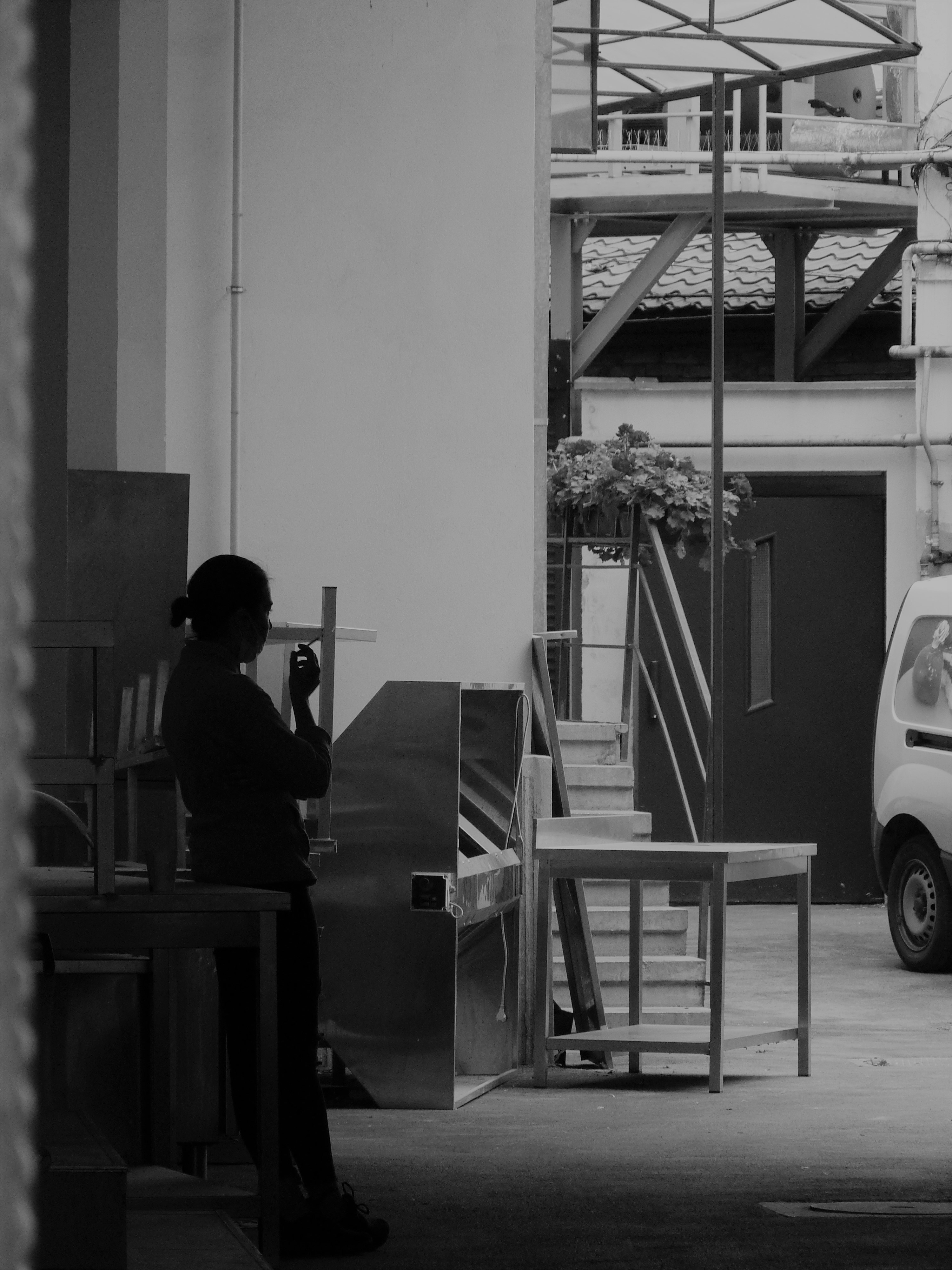 A figure stands in a shadowy corridor, engaged in thought while surrounded by urban elements like furniture and a doorway. The monochrome palette enhances the contemplative mood.