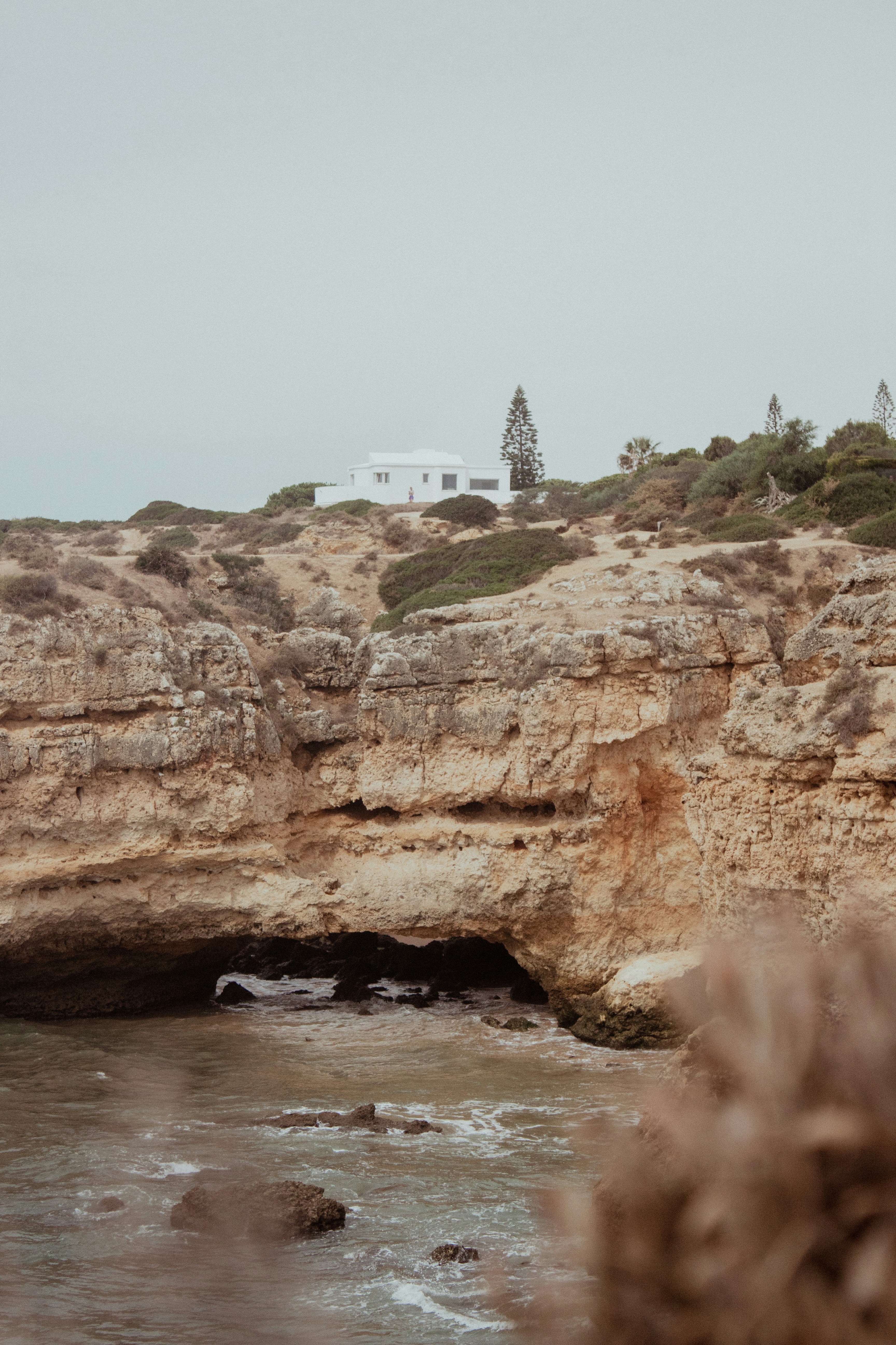 Rocky cliffs with a small cave by the ocean, topped with sparse greenery and distant buildings.