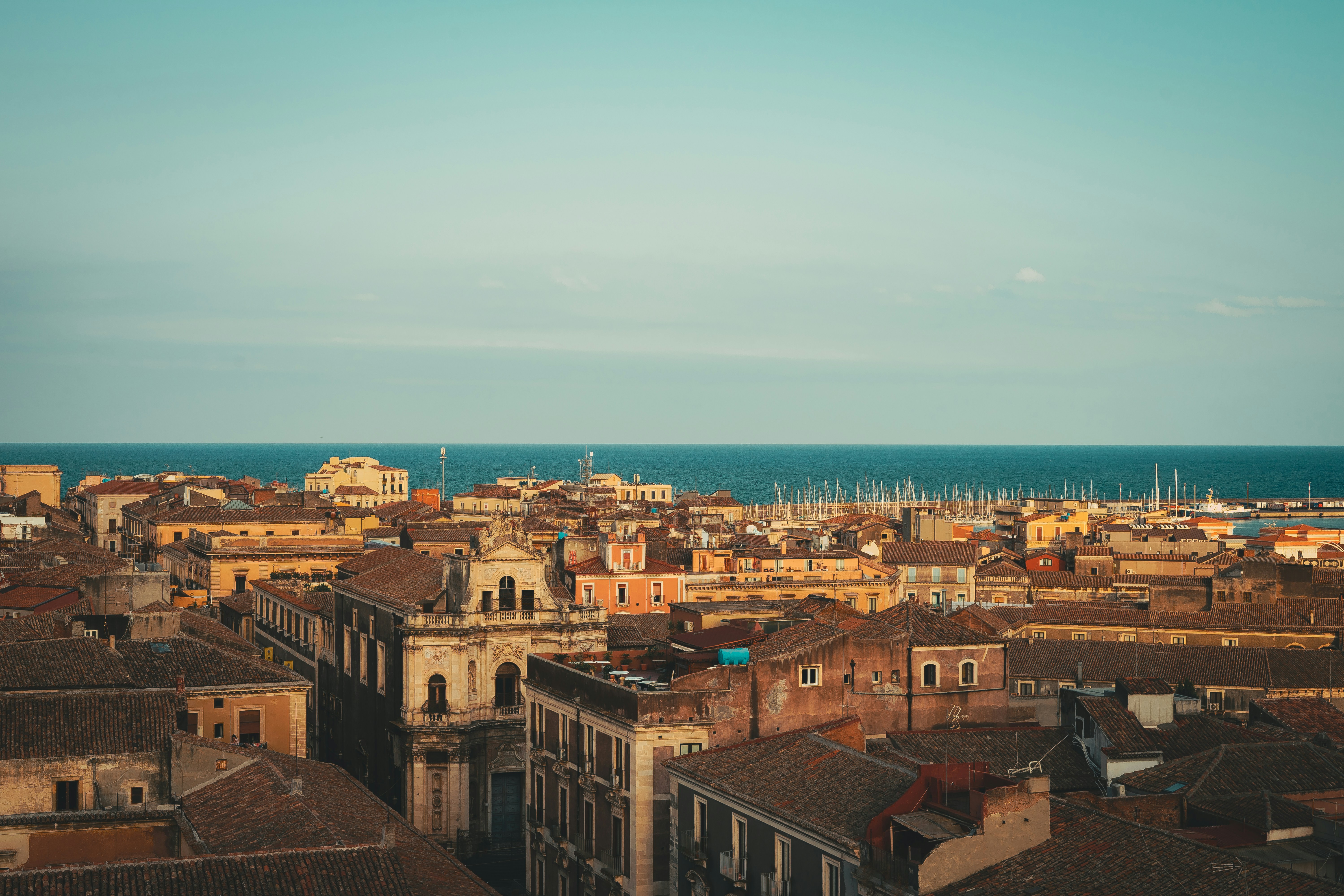 Sunlit cityscape with rustic rooftops and a distant blue sea under a clear sky.
