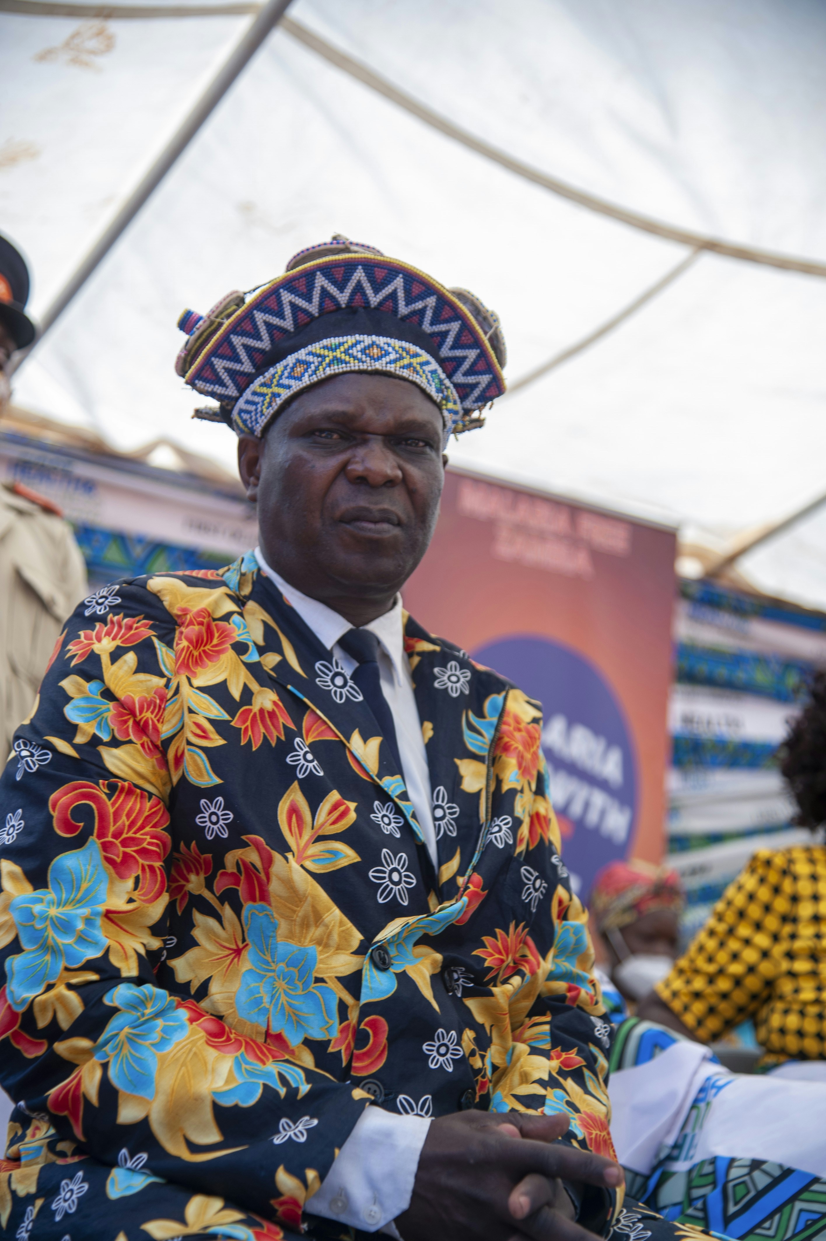 A man in a colorful floral suit and traditional hat seated at a cultural event, embodying local heritage and pride.