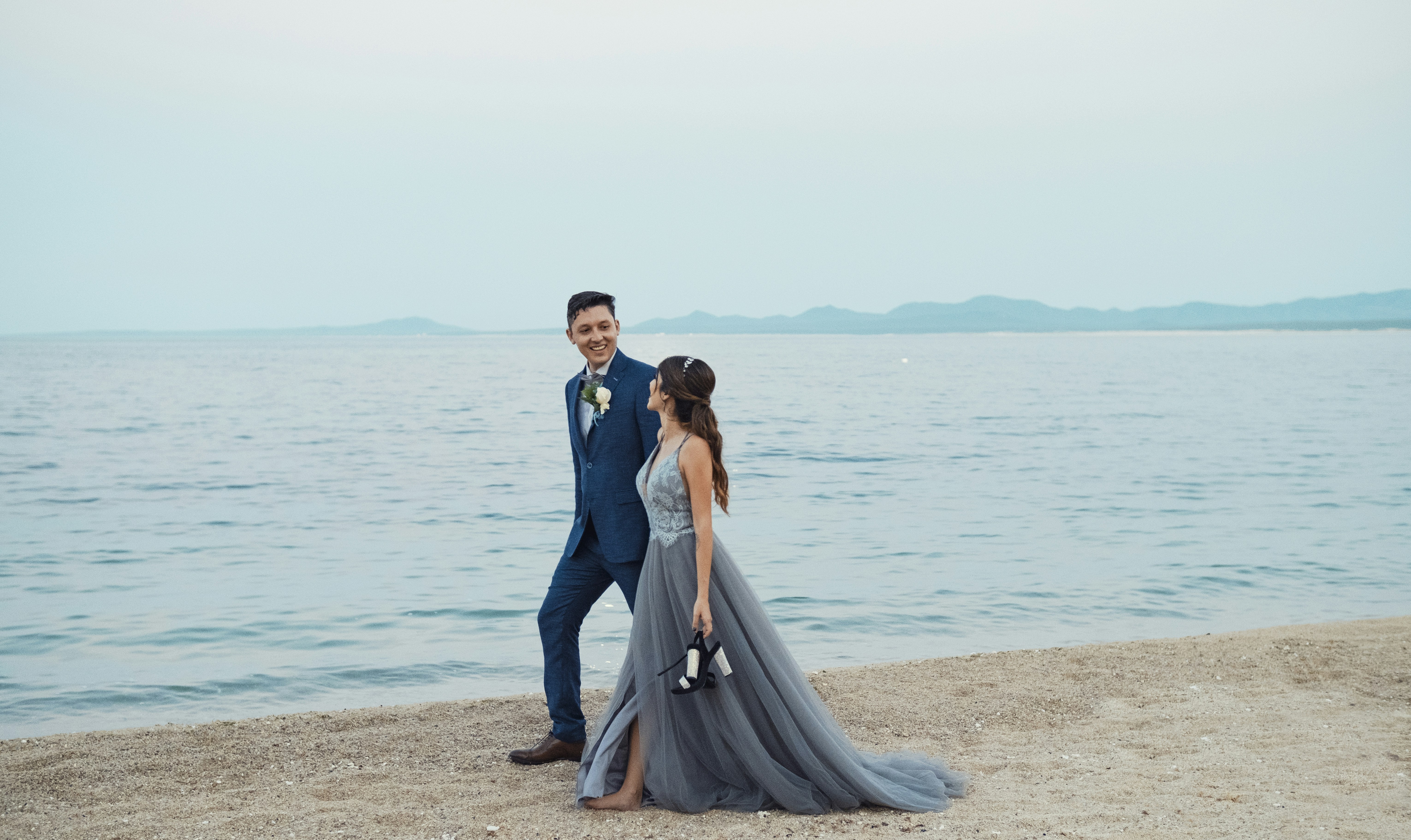 Couple walking along a tranquil beach with distant hills on the horizon.