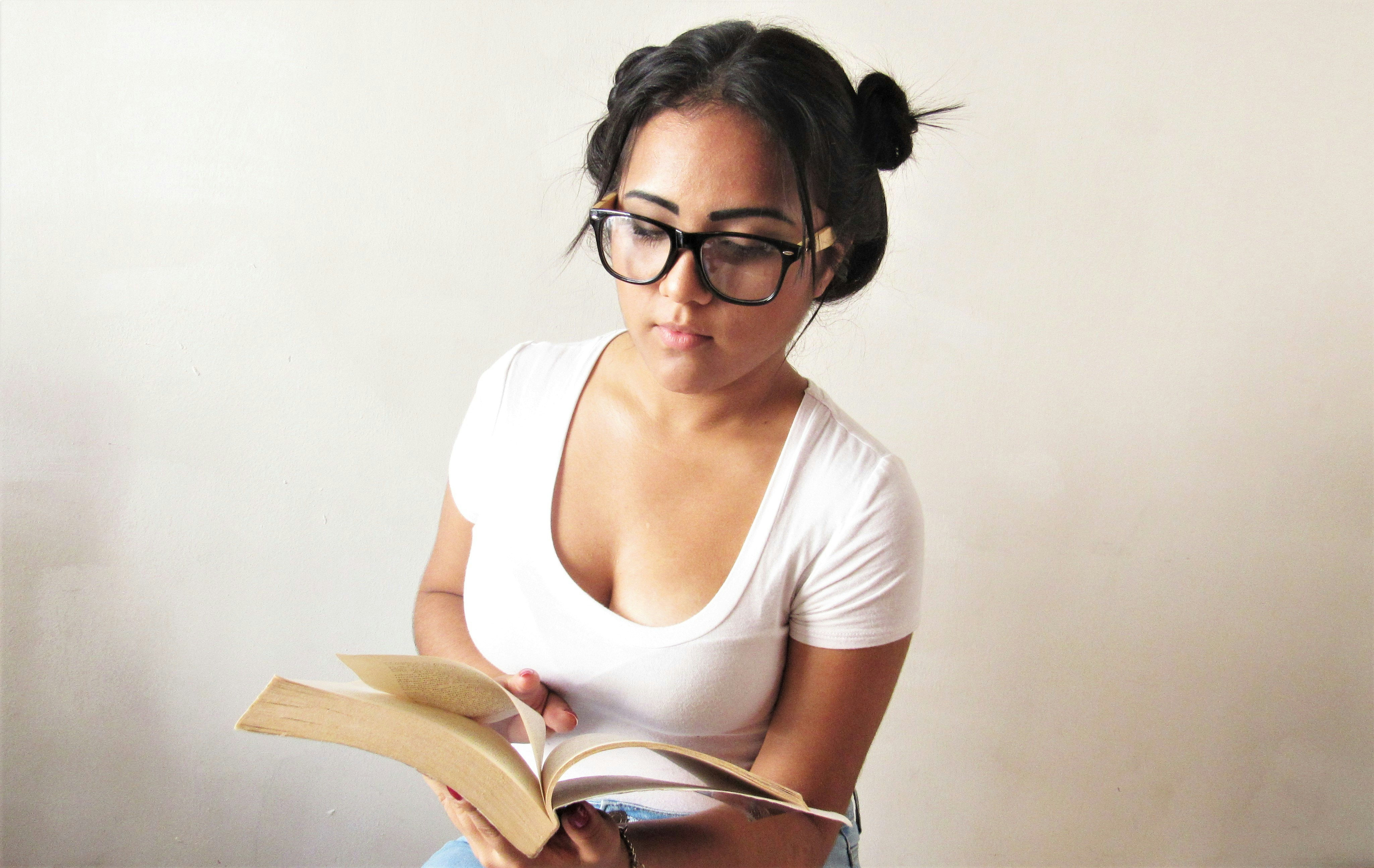 Woman with glasses in a white shirt reading a book against a plain background.