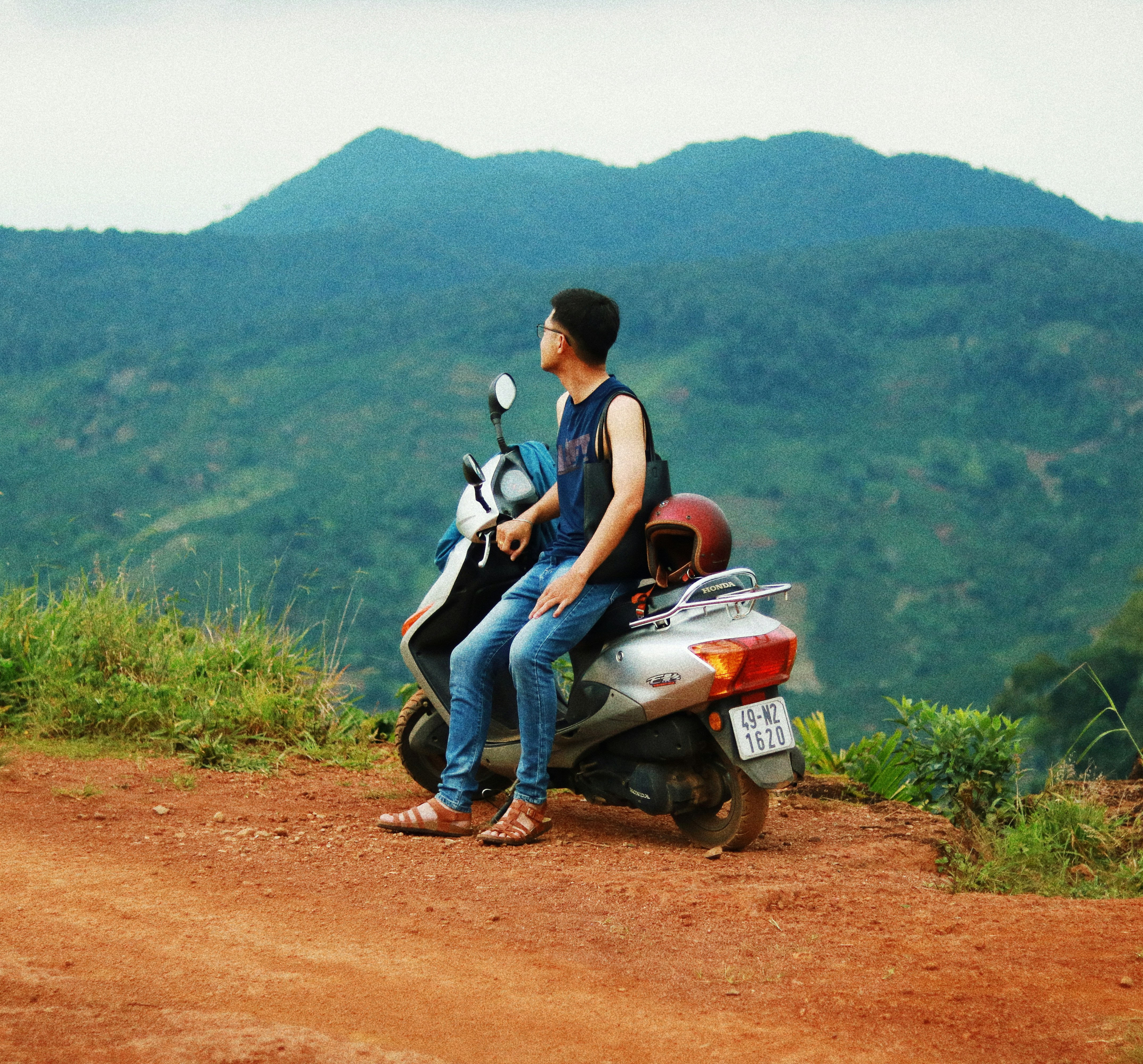 Young man seated on a scooter, gazing at distant mountains under a cloudy sky. The scene captures a tranquil moment in nature.