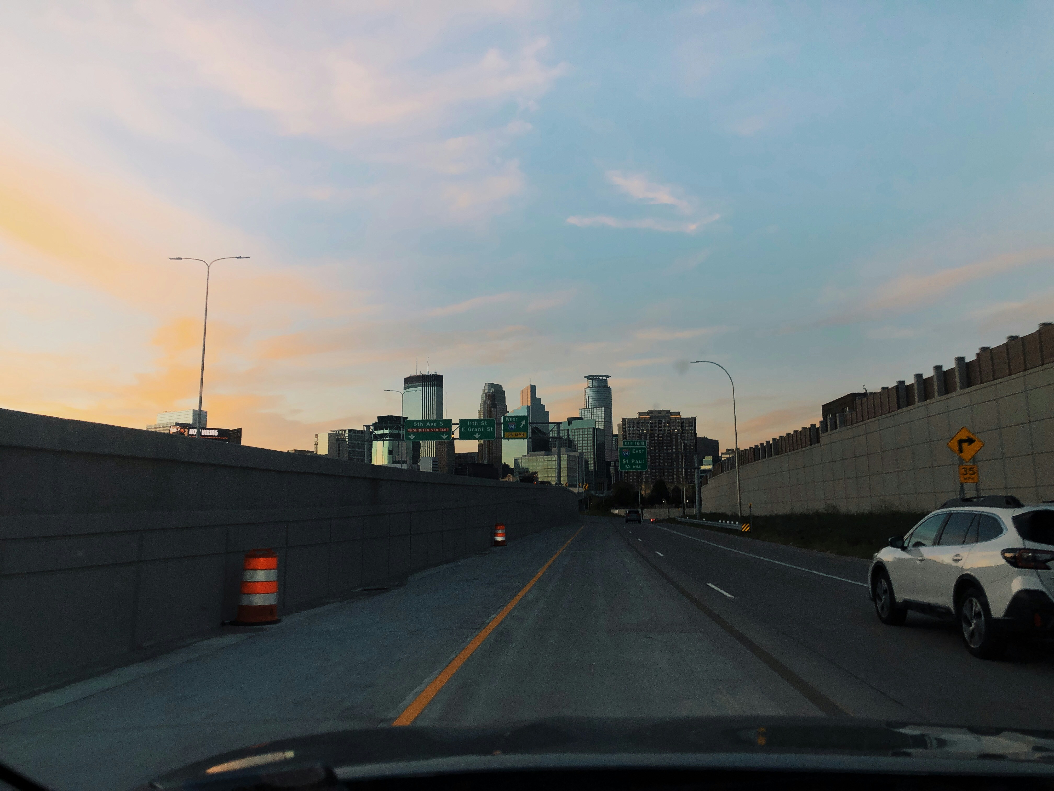 City skyline at dusk viewed from a moving vehicle, showcasing modern architecture and road signs. 