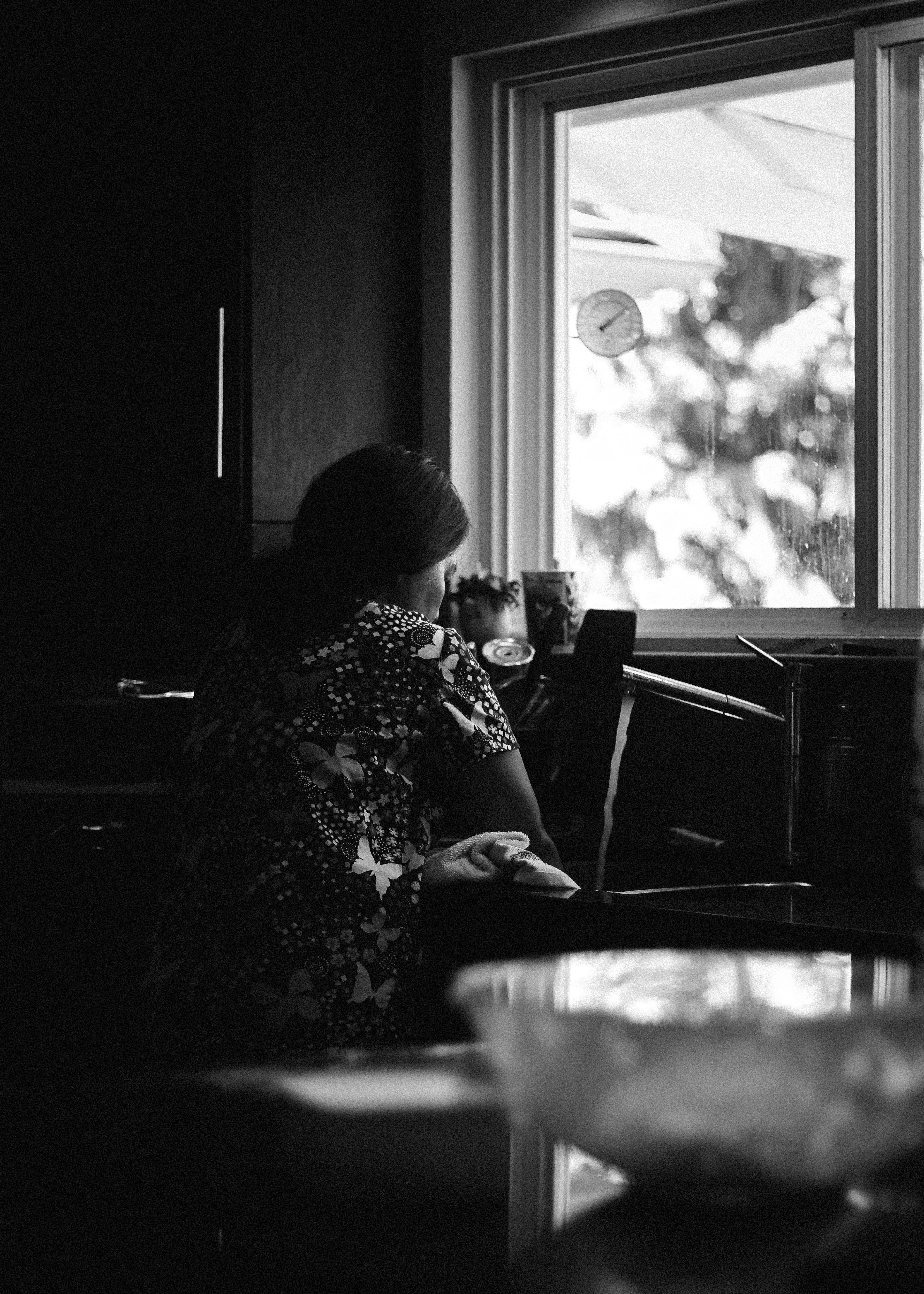 A person with a floral-patterned shirt gazes out a window, framed by dark kitchen elements, evoking a sense of contemplation. The scene captures the interplay of light and shadow.