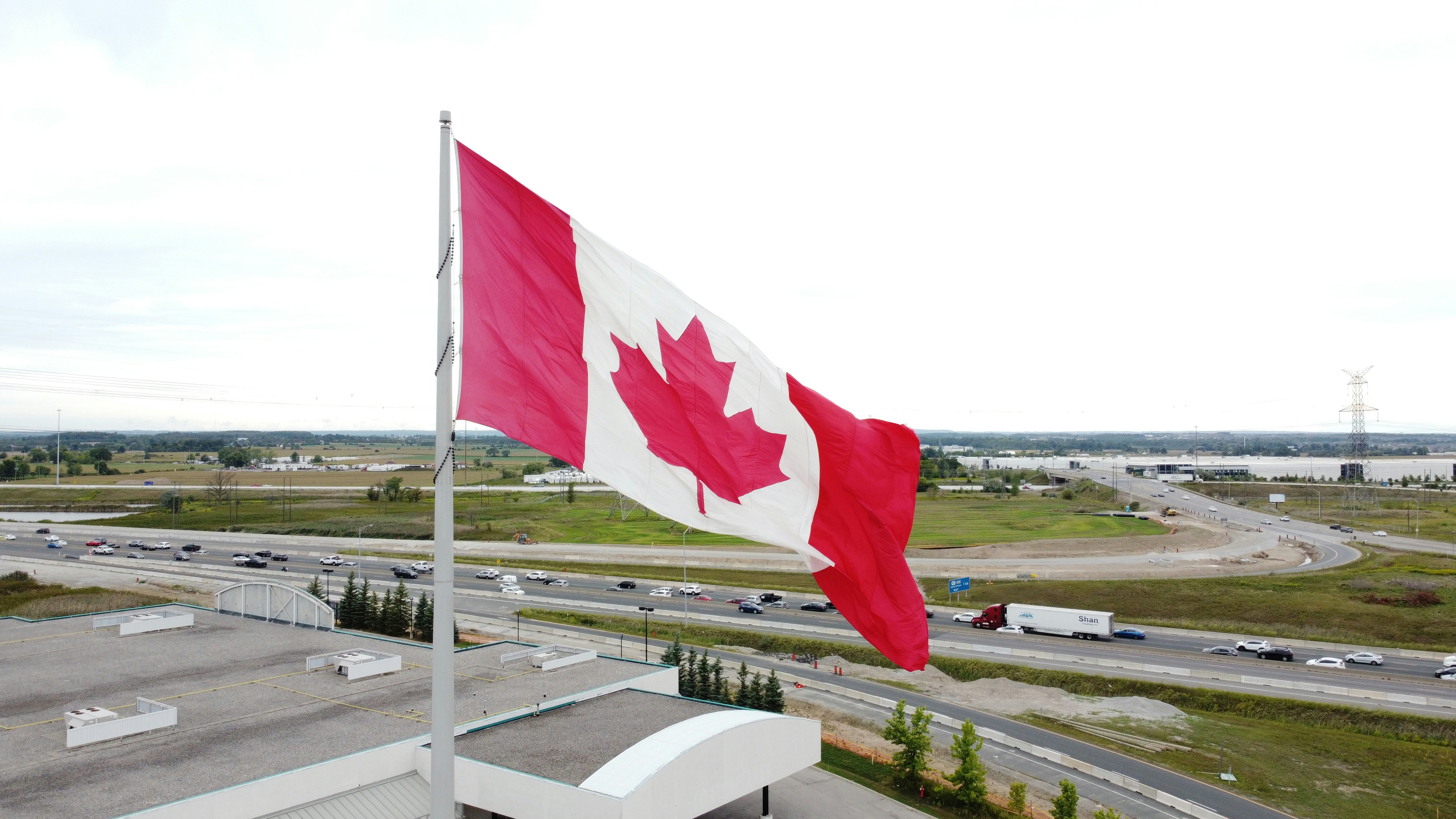Canadian flag billowing in the wind above a busy highway, showcasing national pride against a backdrop of open landscape.