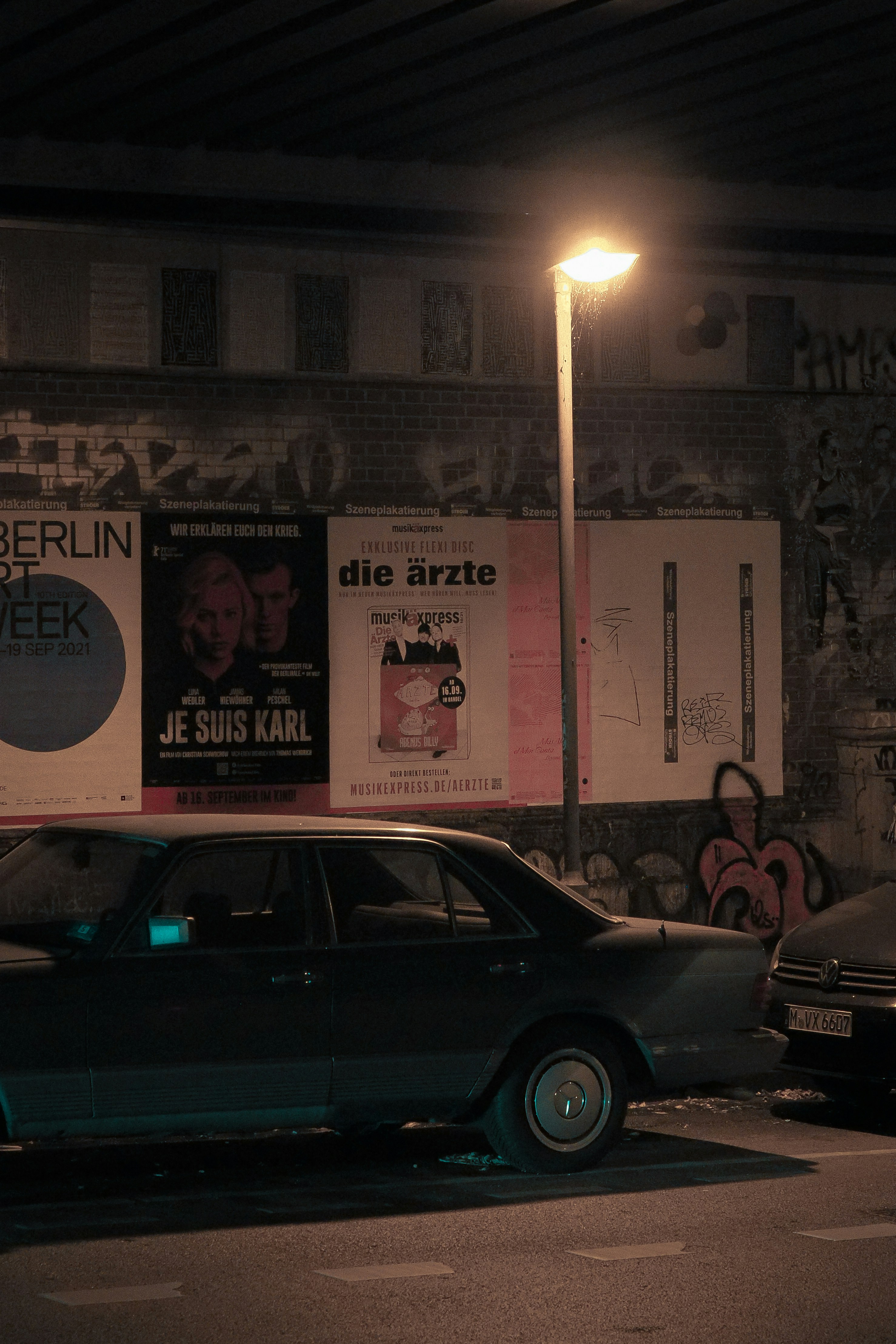 A dimly lit street featuring posters for cultural events, illuminated by a streetlight, with parked cars in the foreground.