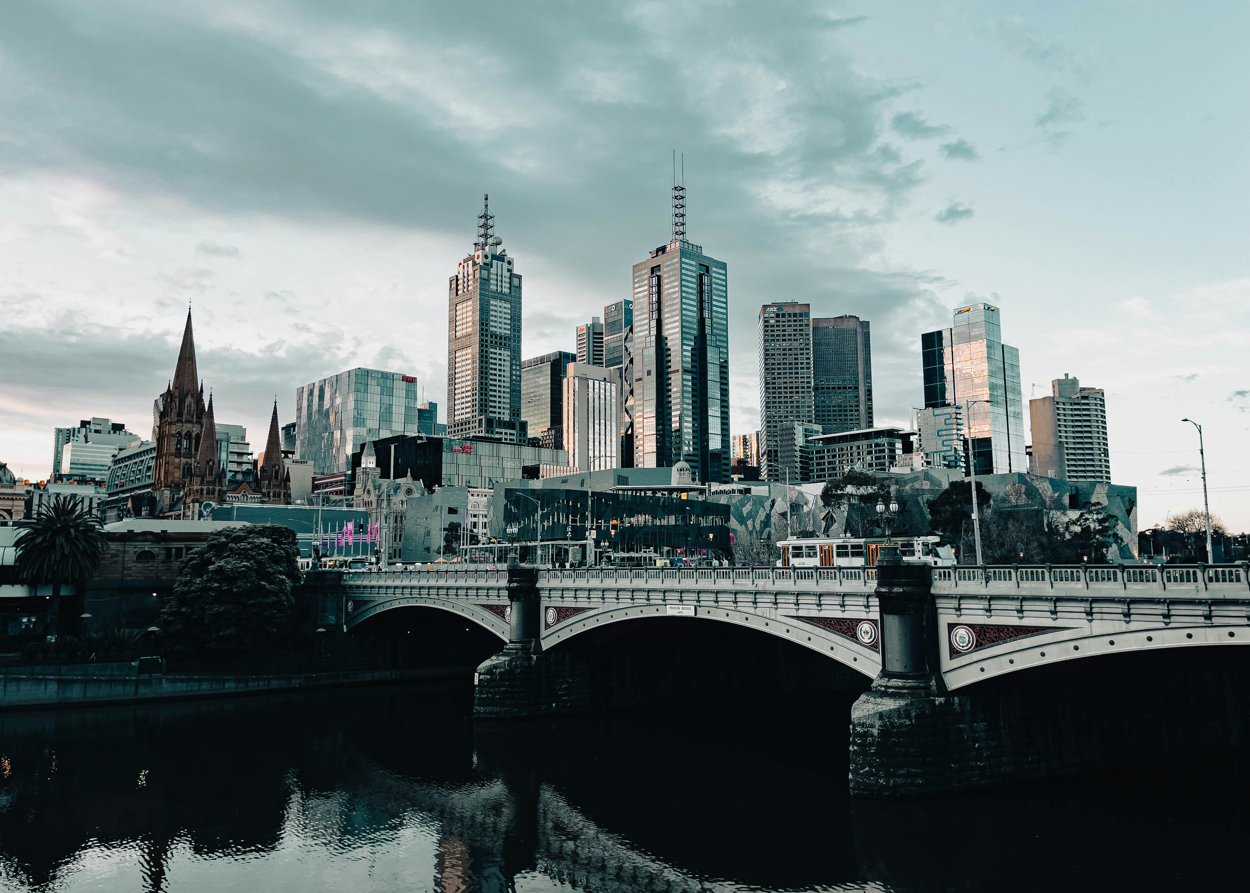 Bridge spanning the Yarra River with Melbourne's skyline featuring iconic skyscrapers under a cloudy sky.