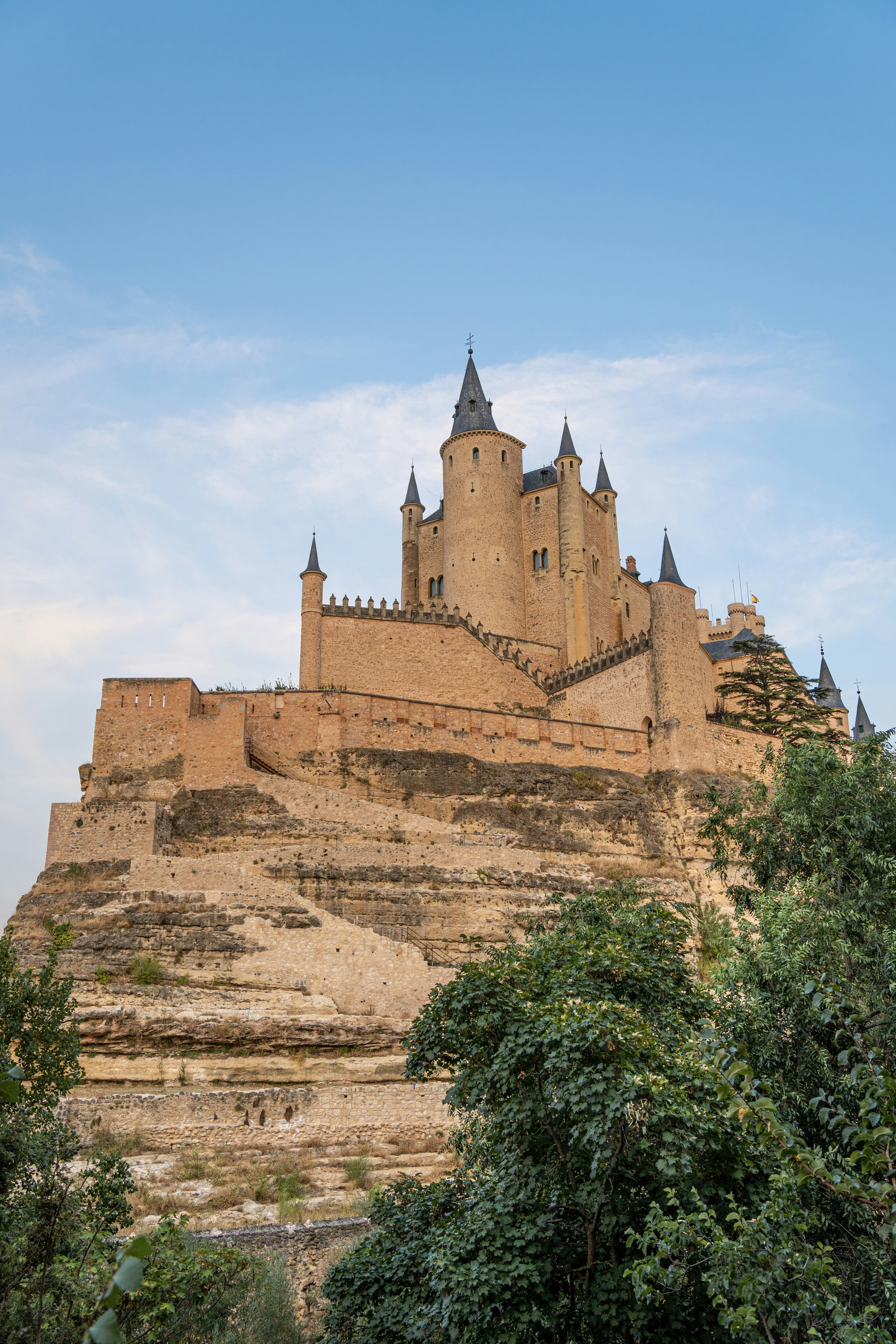 Castle perched atop a rocky hill surrounded by lush greenery and a clear blue sky.