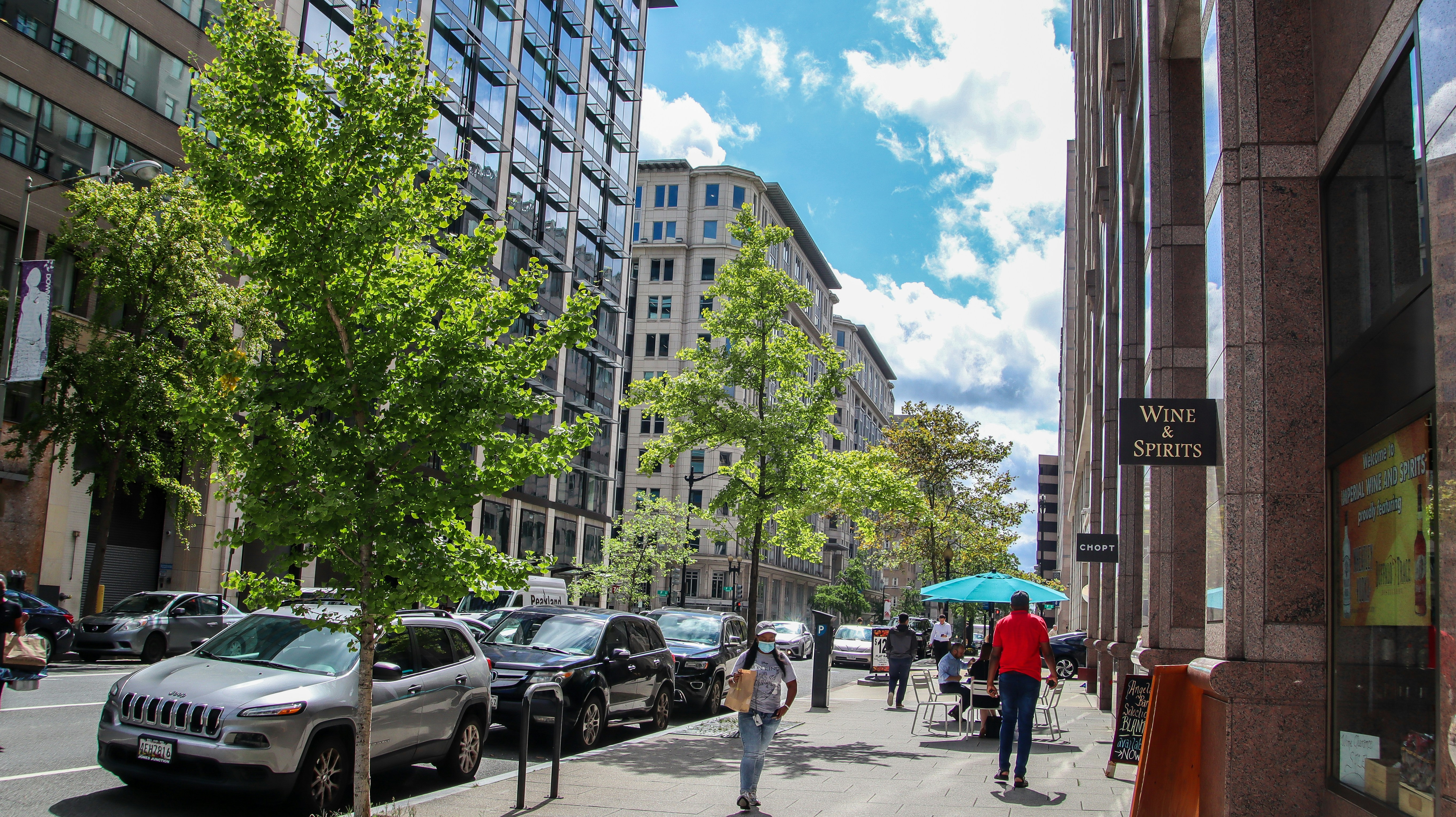 Bustling city street lined with trees and parked cars, featuring pedestrians enjoying a sunny day. A wine and spirits shop is visible on the right.