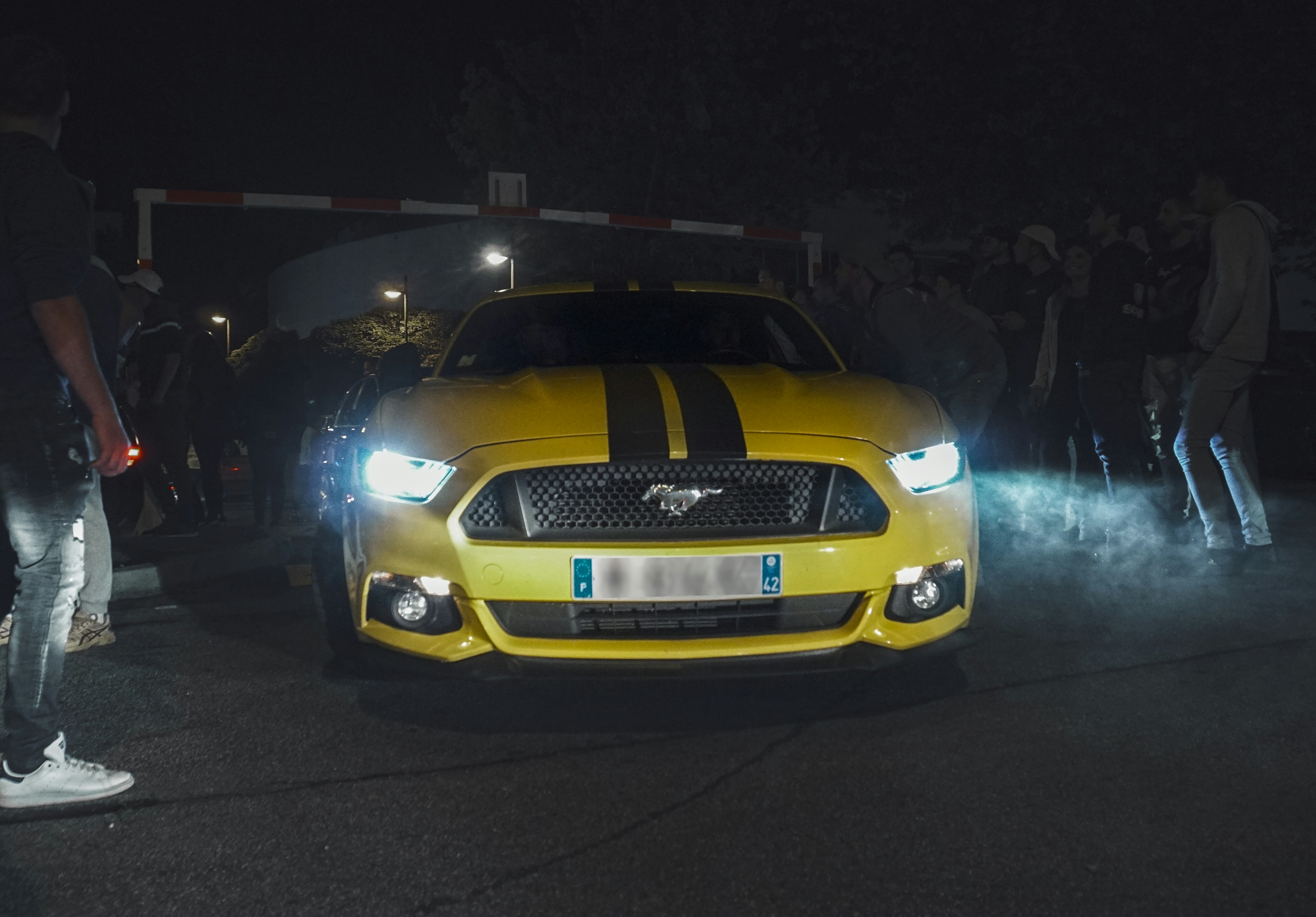 Yellow sports car with black stripes illuminated by headlights in a dimly lit street scene.