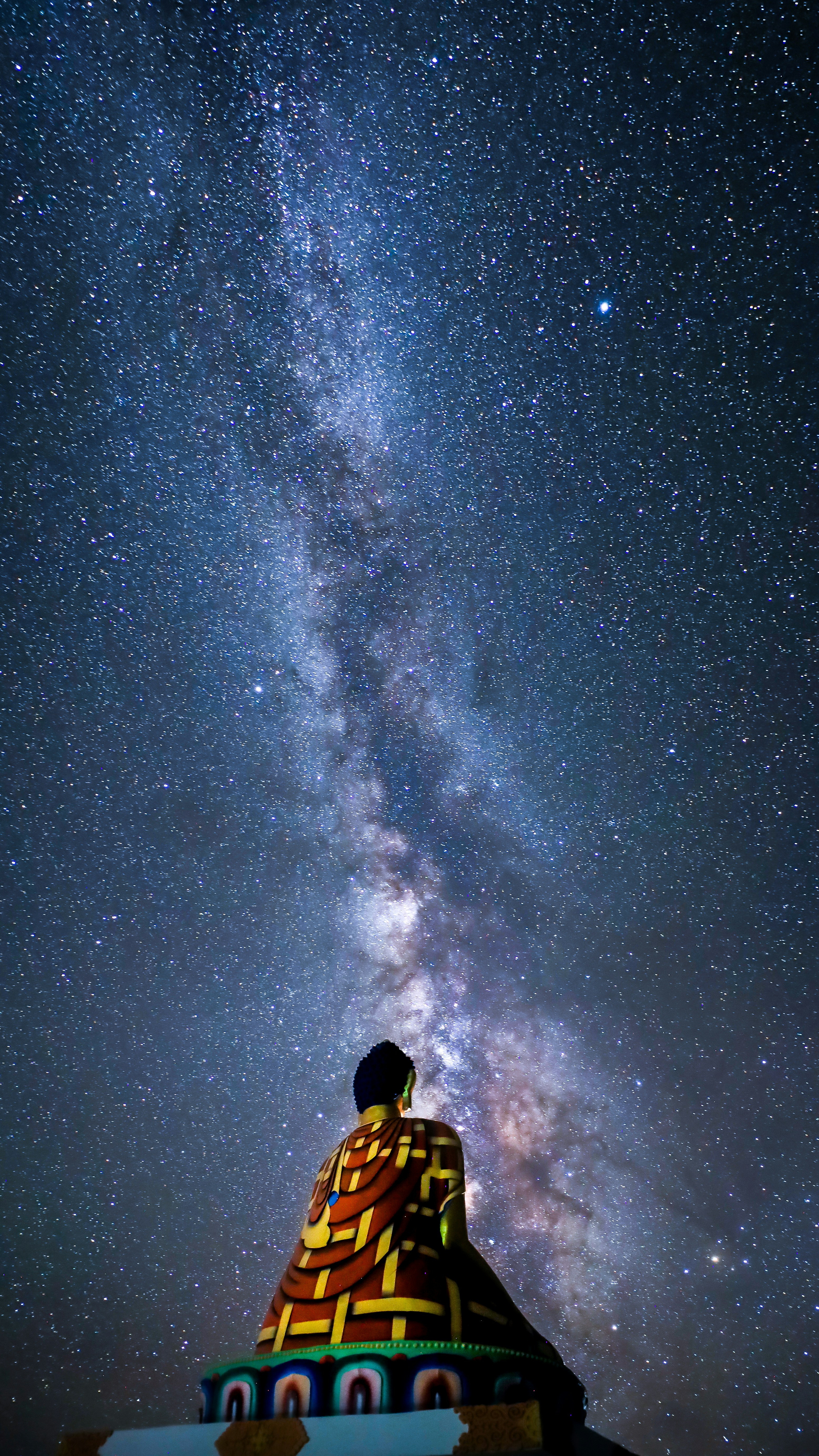 A colorful statue of a seated figure gazes up at the expansive Milky Way, surrounded by countless stars in a clear night sky.