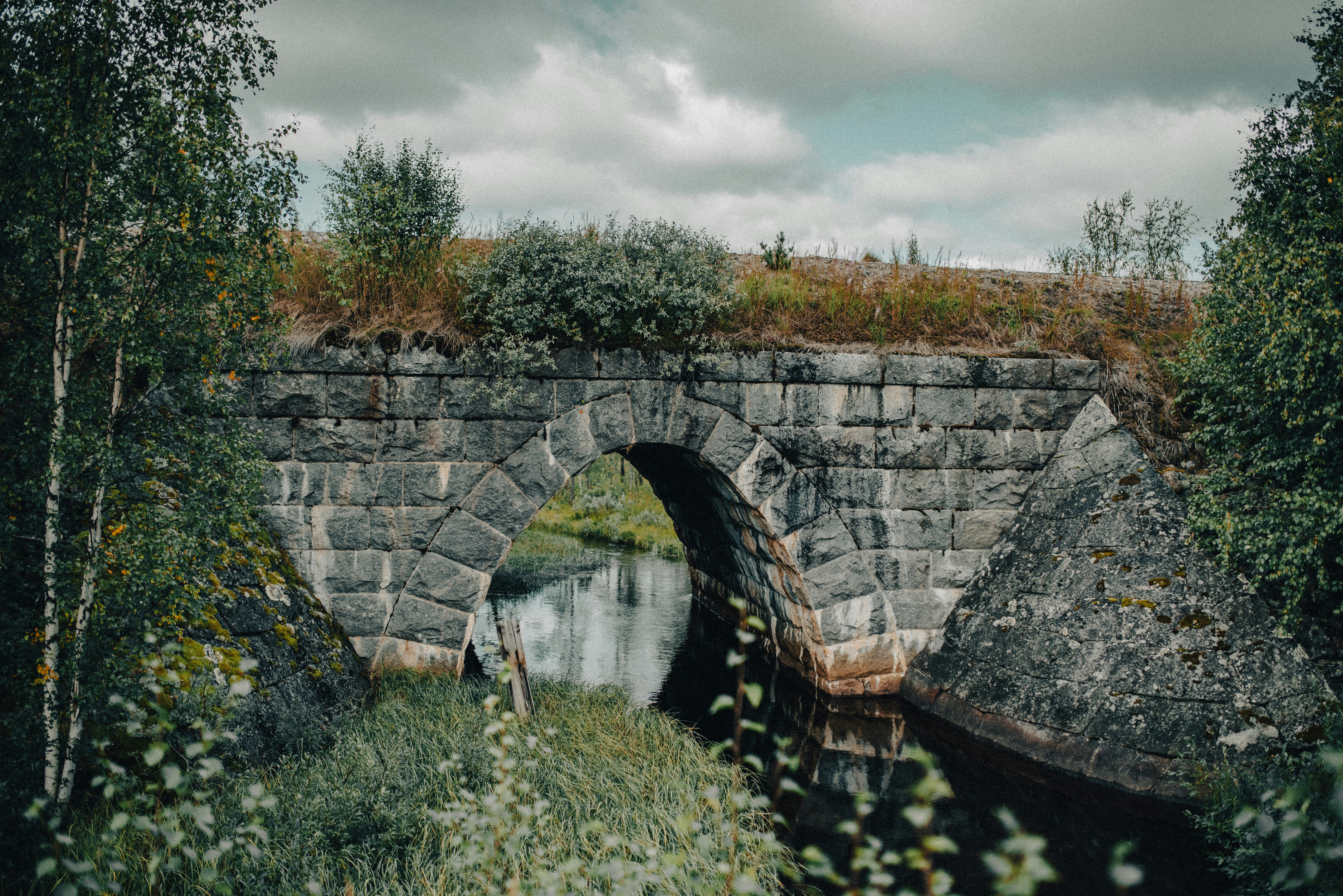 Ancient stone arch bridge spanning a serene waterway, surrounded by lush greenery and soft clouds above.