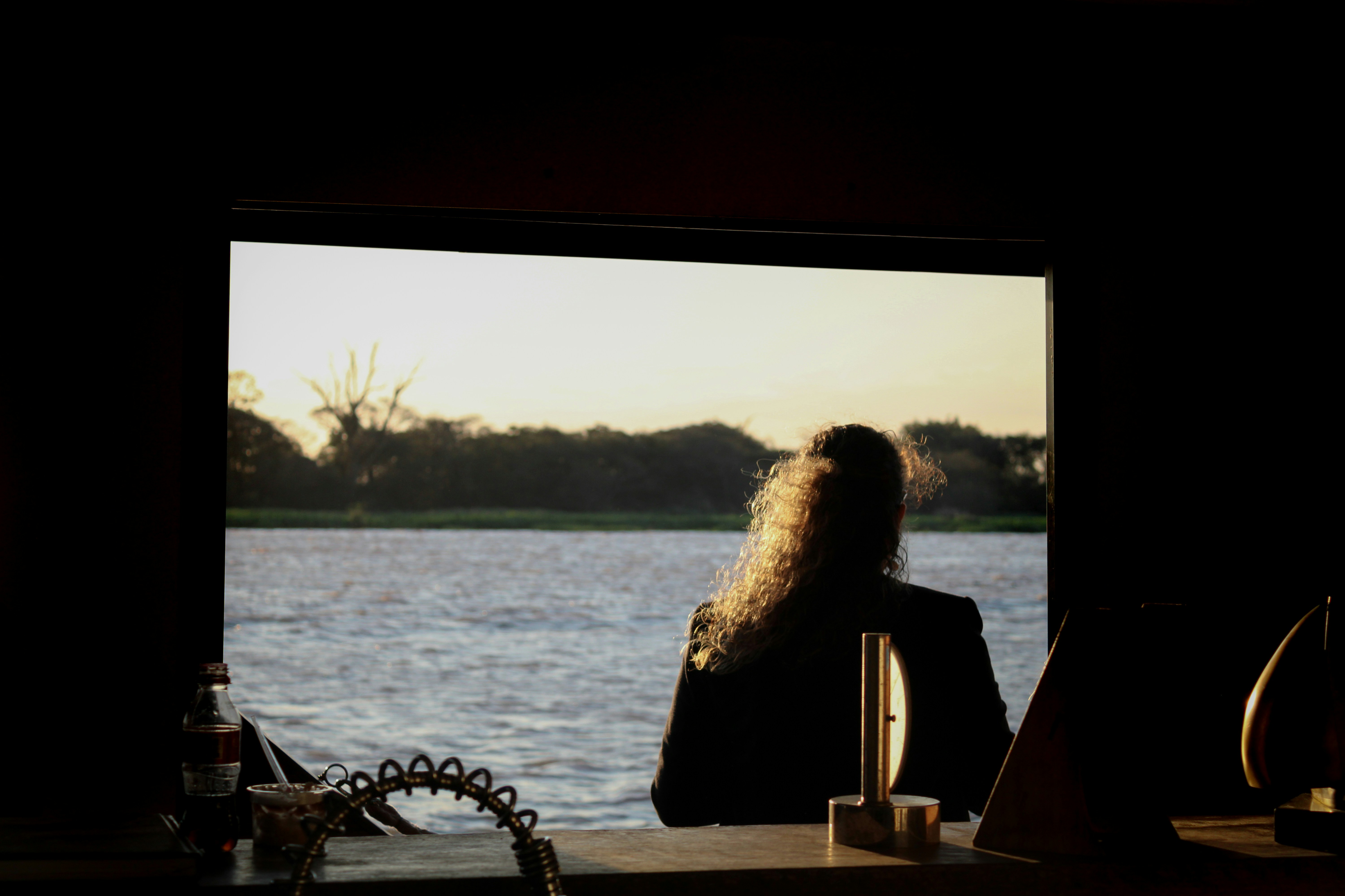 Silhouette of a person gazing out of a window towards a tranquil river at sunset, framed by the interior of a boat.
