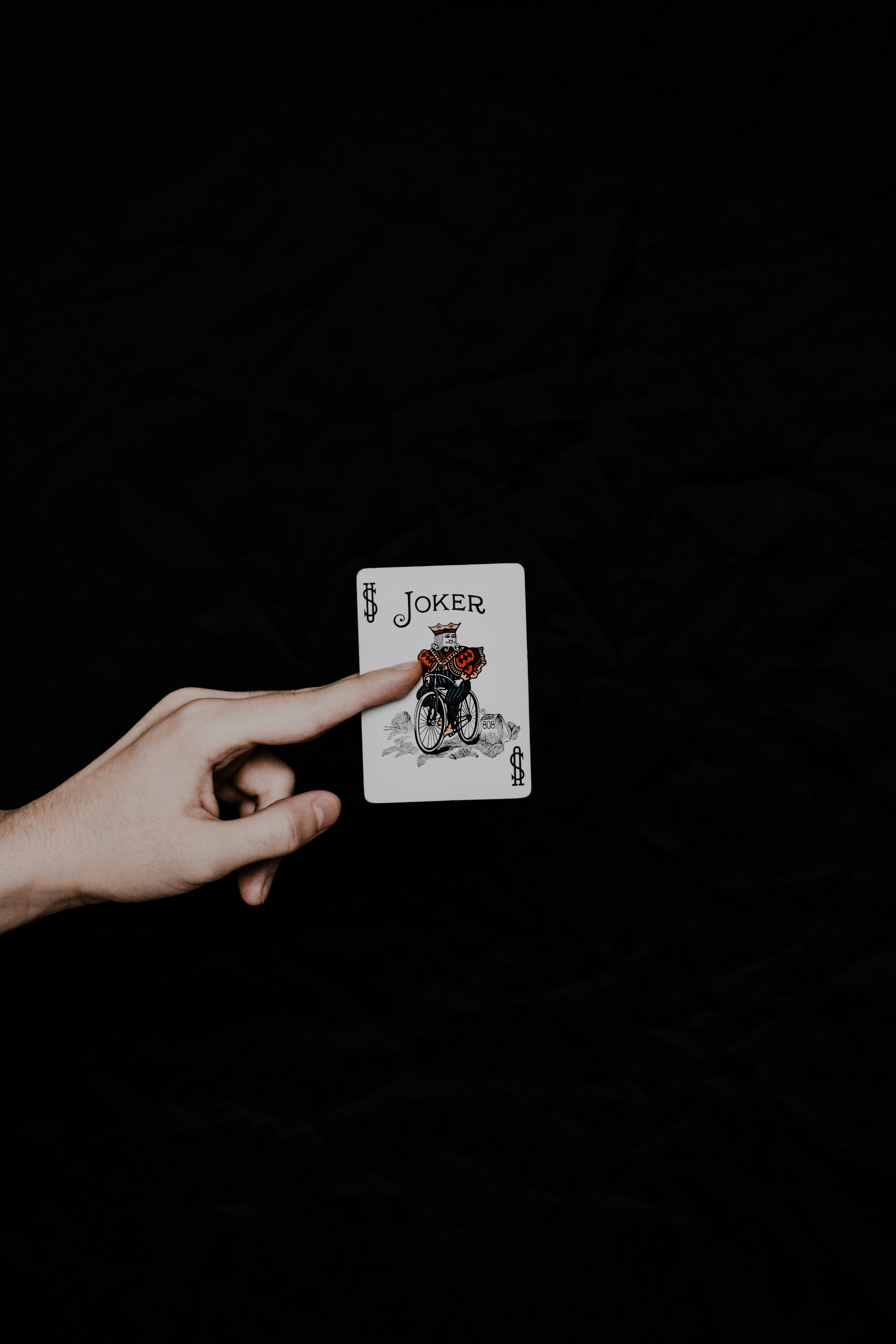 A hand delicately points to a Joker playing card against a dark background, emphasizing its playful design and intricate details.
