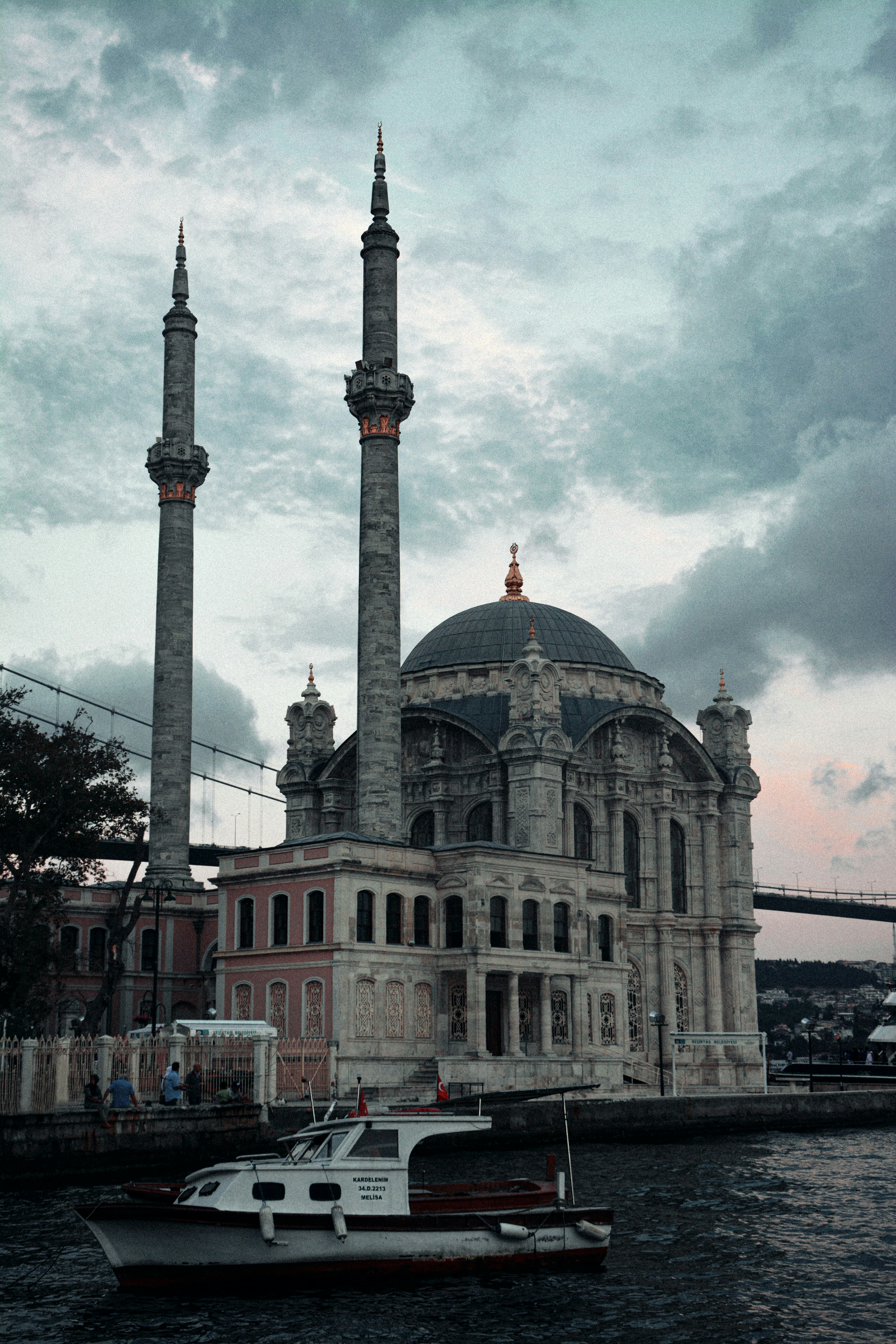 Historic waterfront mosque with twin minarets, framed by a serene river and a small boat. The sky is adorned with soft clouds, hinting at the evening light.