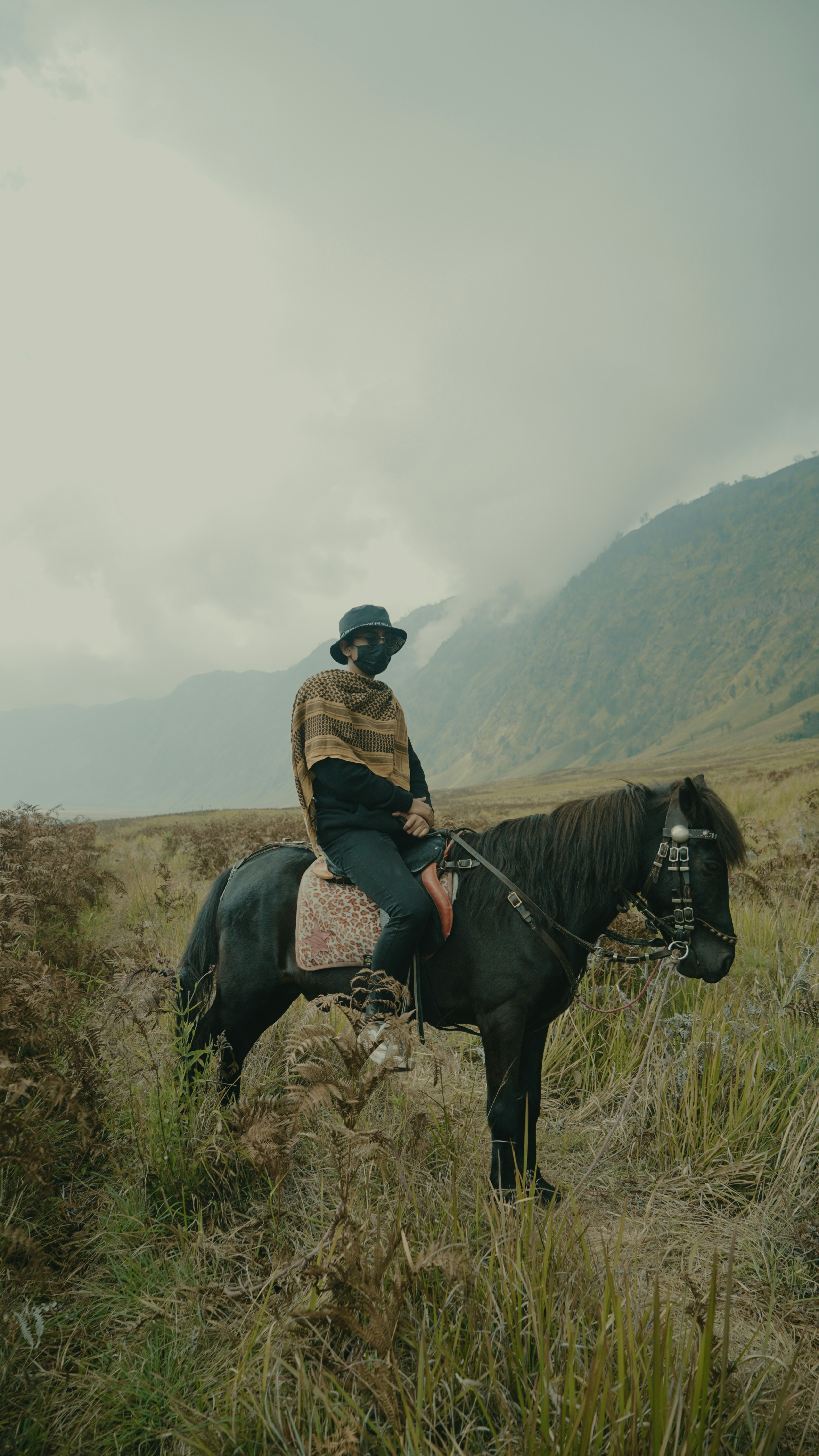 A lone rider on a dark horse stands in a misty hillside field, wearing a patterned shawl and wide-brimmed hat; the scene is calm and expansive, with mountains fading into clouds behind. This is a photographic capture of a western-style landscape with a solitary figure.