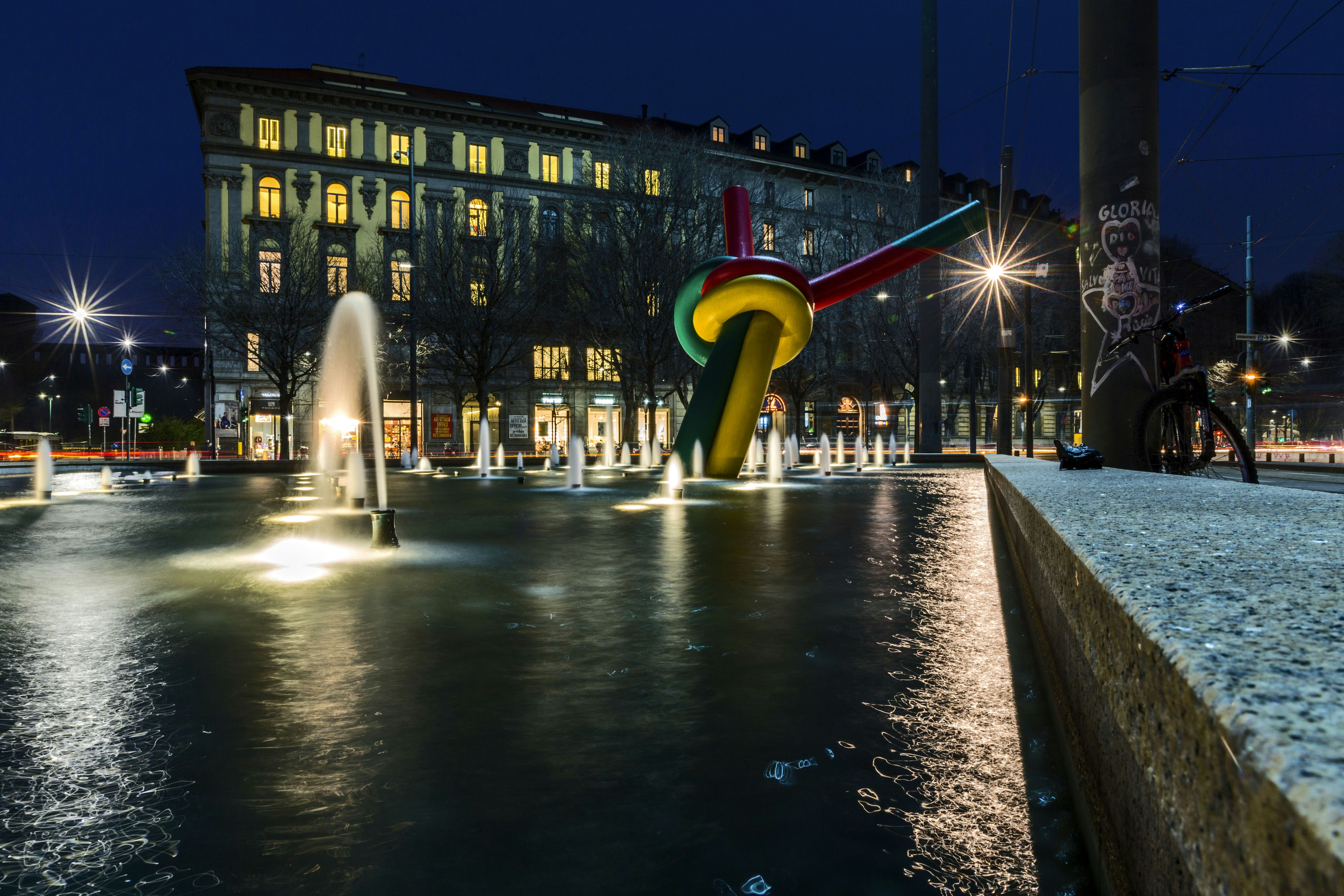 Illuminated fountains and a vibrant sculpture enliven a city square at night.