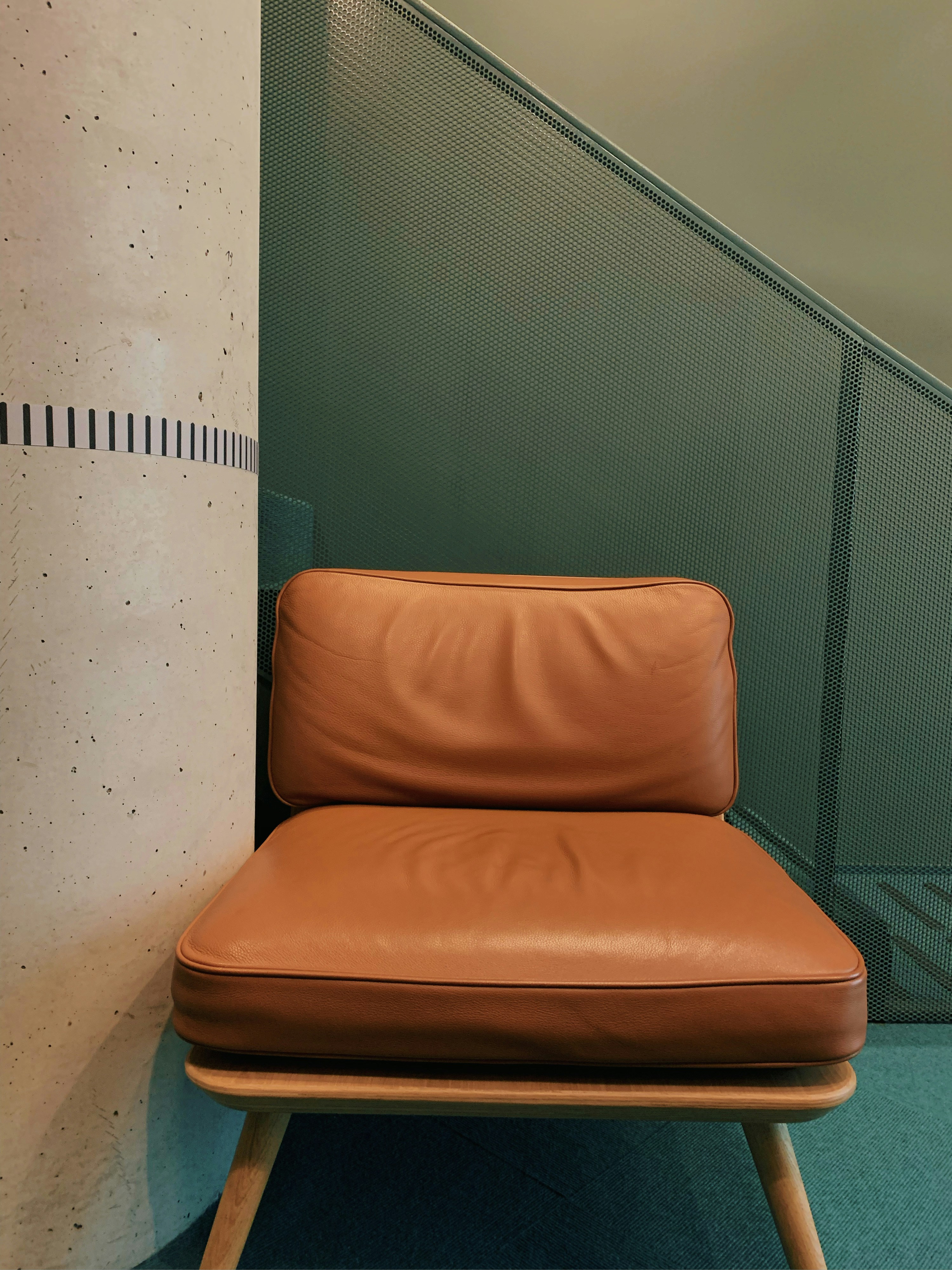 A cozy brown leather chair positioned next to a textured column, with a backdrop of green stairs. The design emphasizes simplicity and comfort.