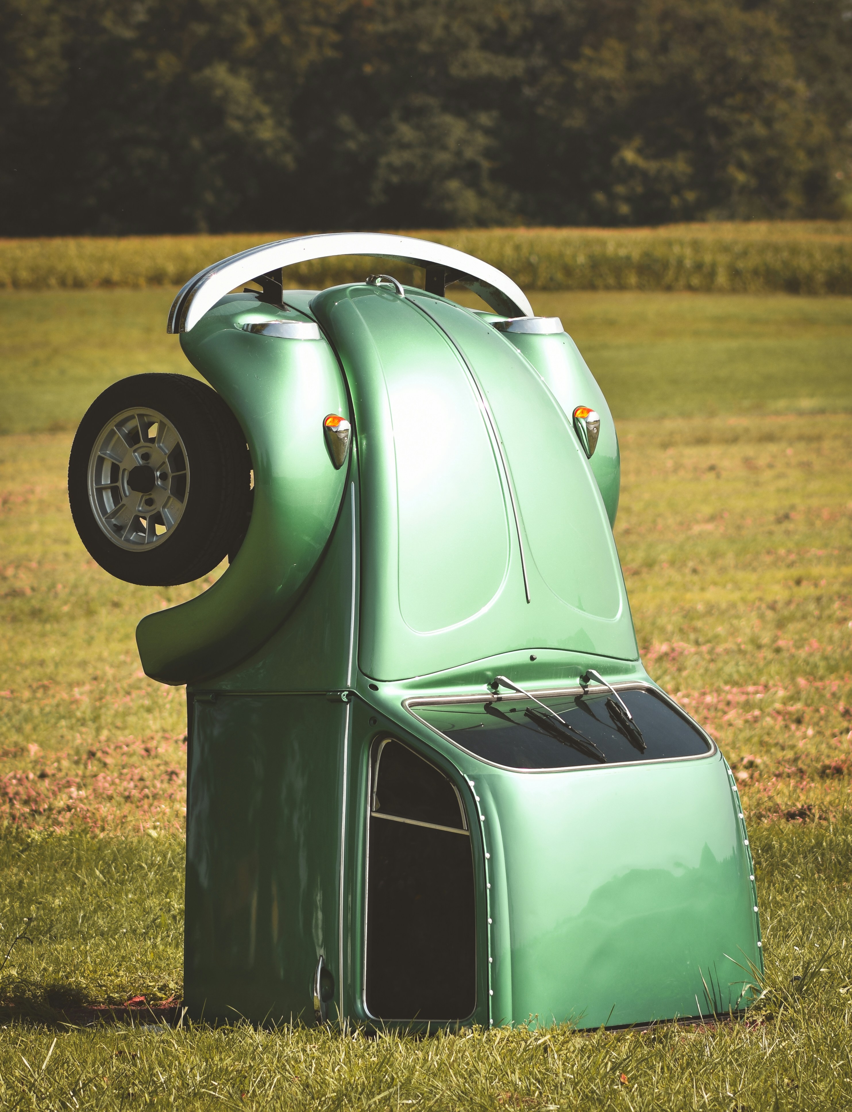 A creatively designed sculpture resembling a vintage car turned upside down, set against a grassy field backdrop.